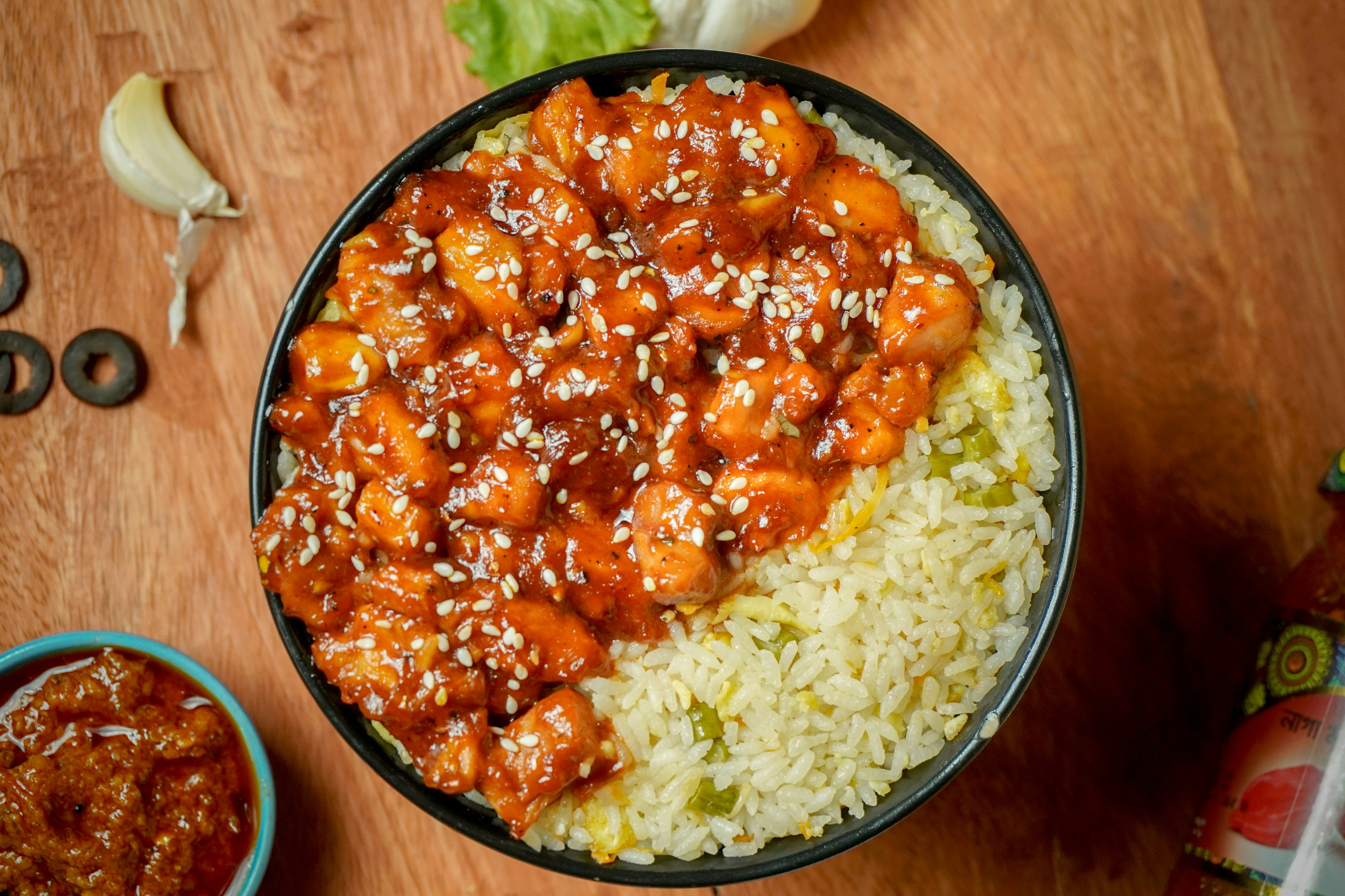 A delicious bowl of glazed chicken with sesame seeds served over seasoned fried rice, accompanied by spicy sauce and garlic cloves on wooden table.