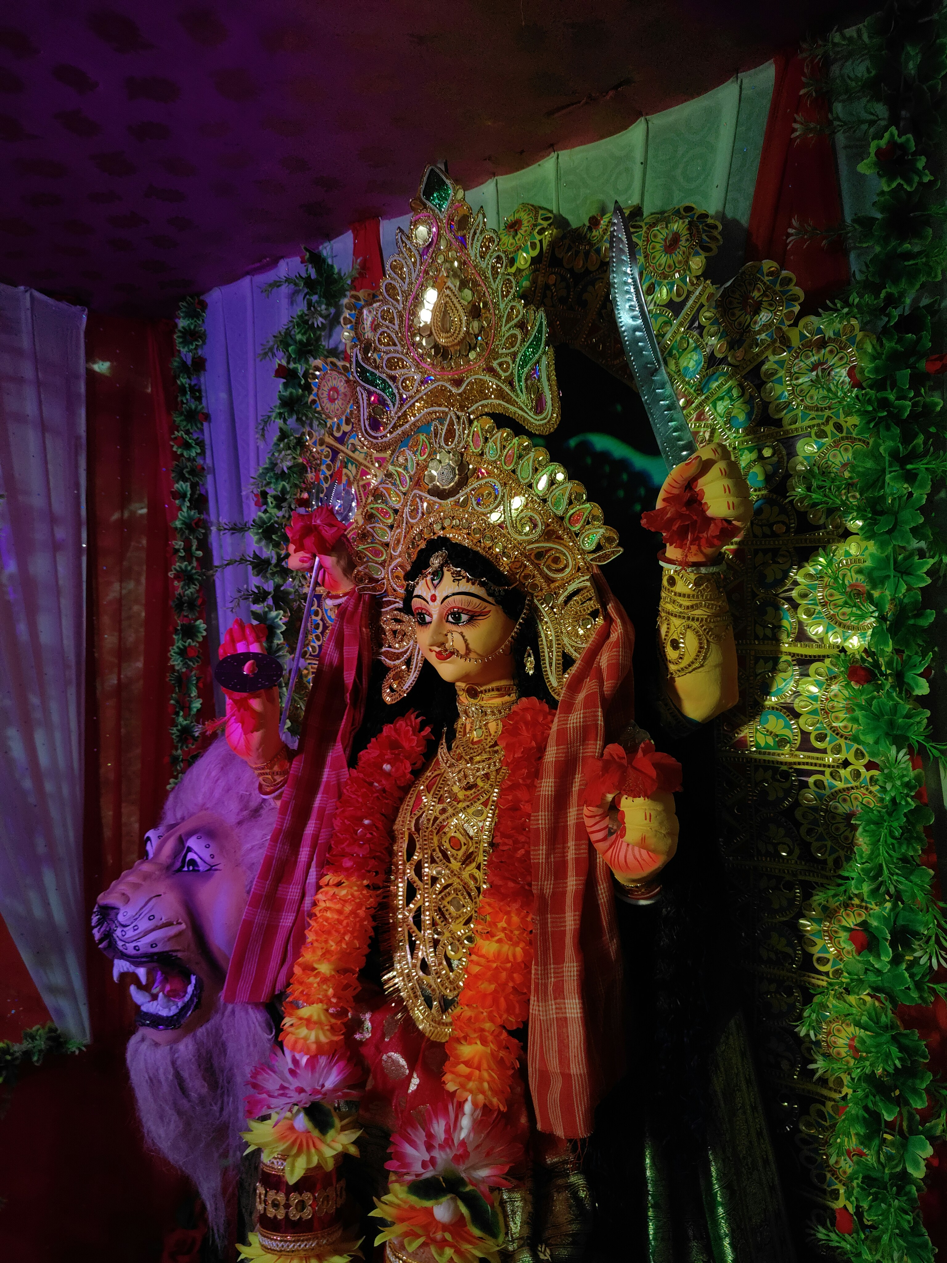 Decorative deity statue adorned with gold jewelry and a jeweled headdress in a festive setup with colorful drapery and artificial greenery.