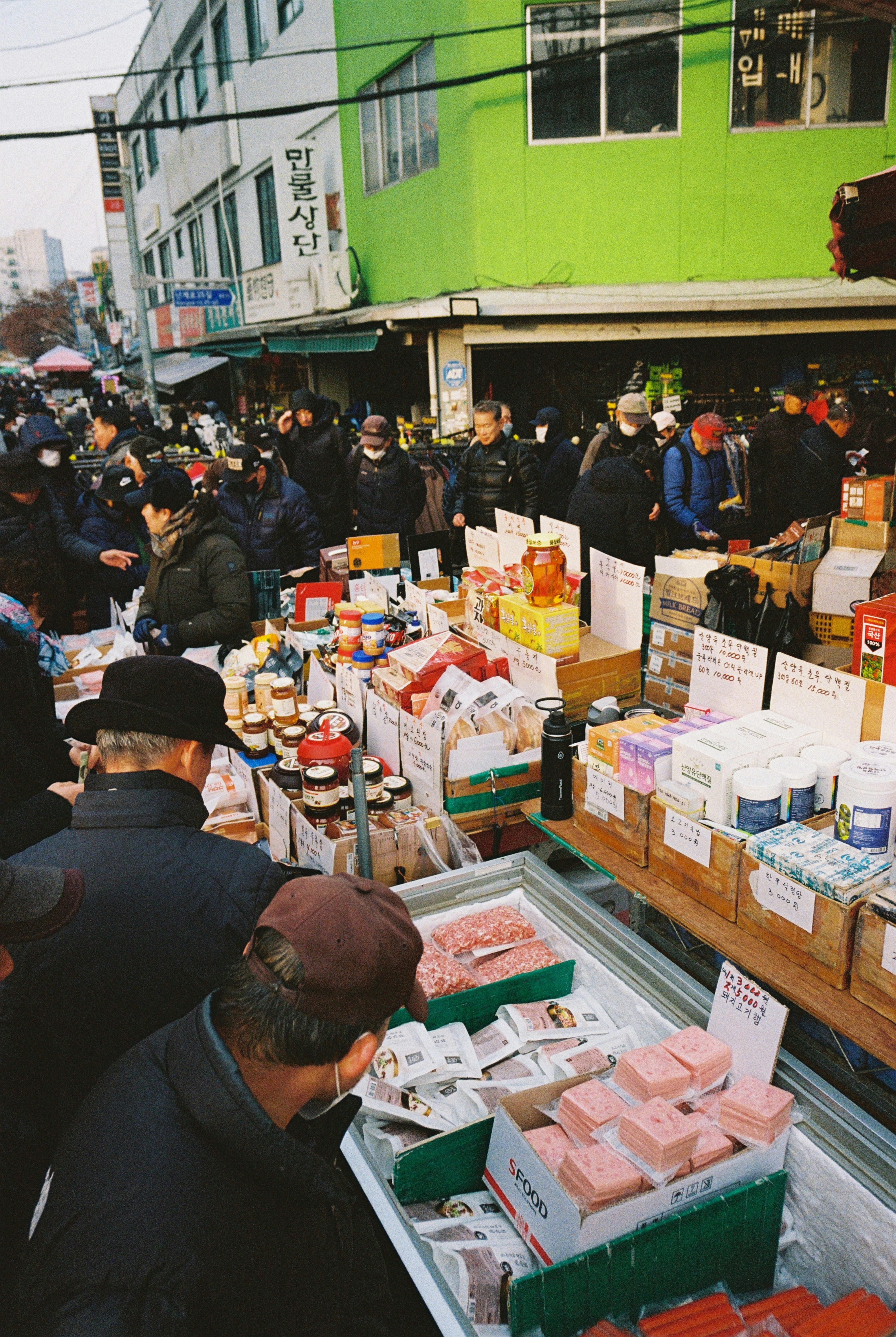Colorful street market scene with customers browsing stalls of meats, packaged goods, and beverages; lively crowd under bright storefronts in the background. This is a documentary-style capture of a busy market moment.