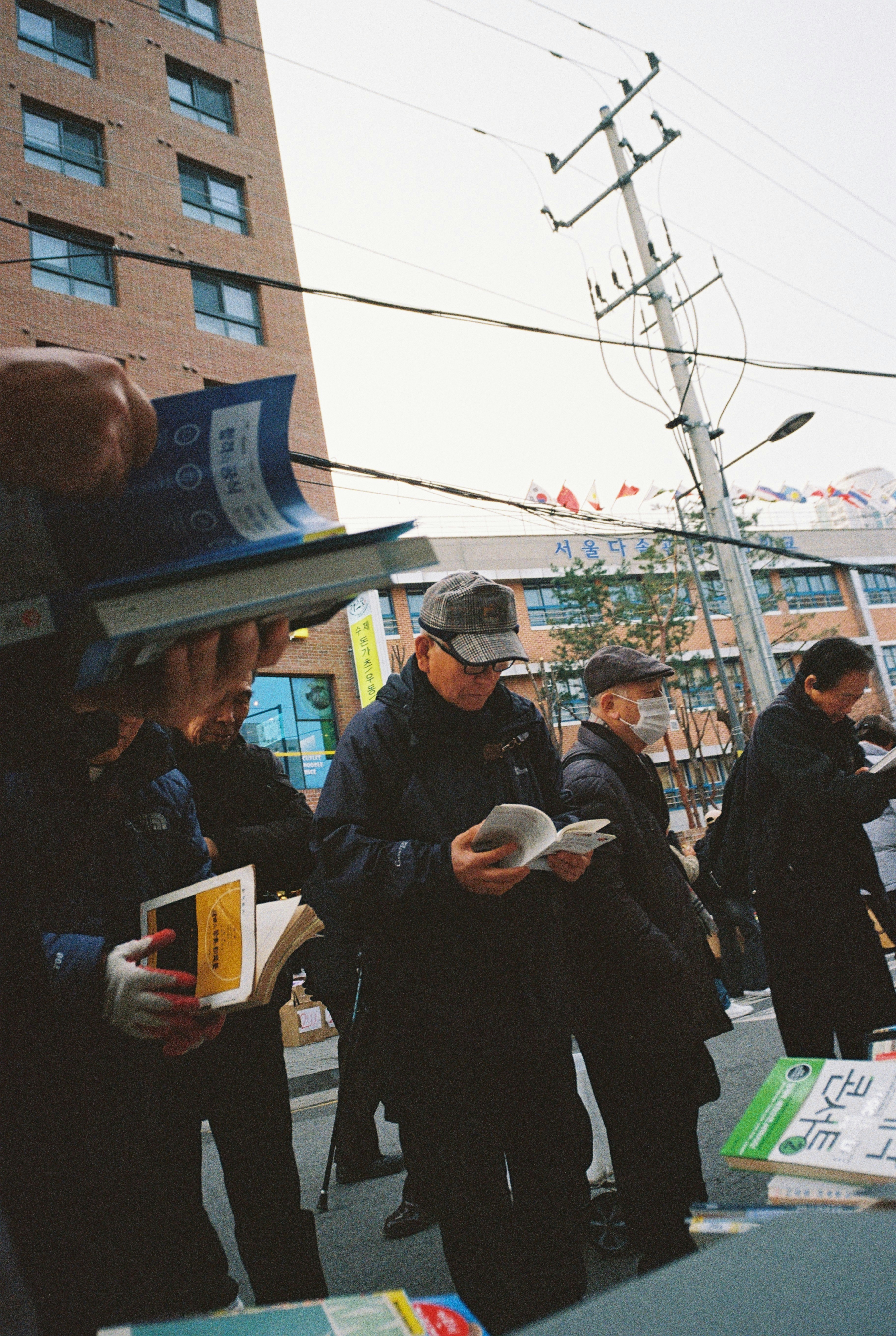 A street-side scene shows several people reading books, with a man in a cap at the center engrossed in a page. The foreground stacks of books hint at a pop-up book market or book fair.