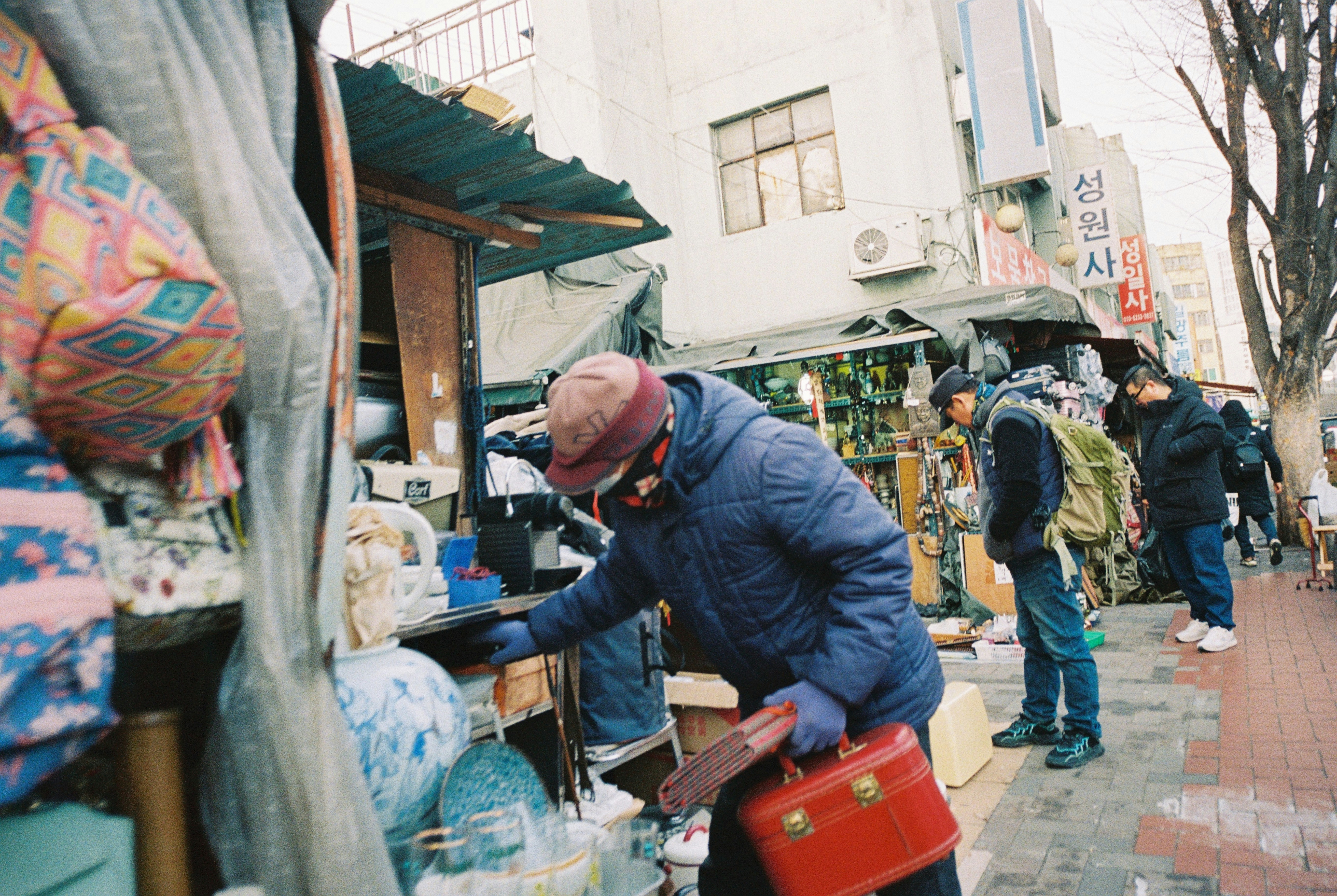 Vibrant street market scene with a blue-jacketed shopper examining items at a stall; pedestrians and storefronts line a narrow sidewalk. A candid snapshot capturing daily commerce in an urban setting.