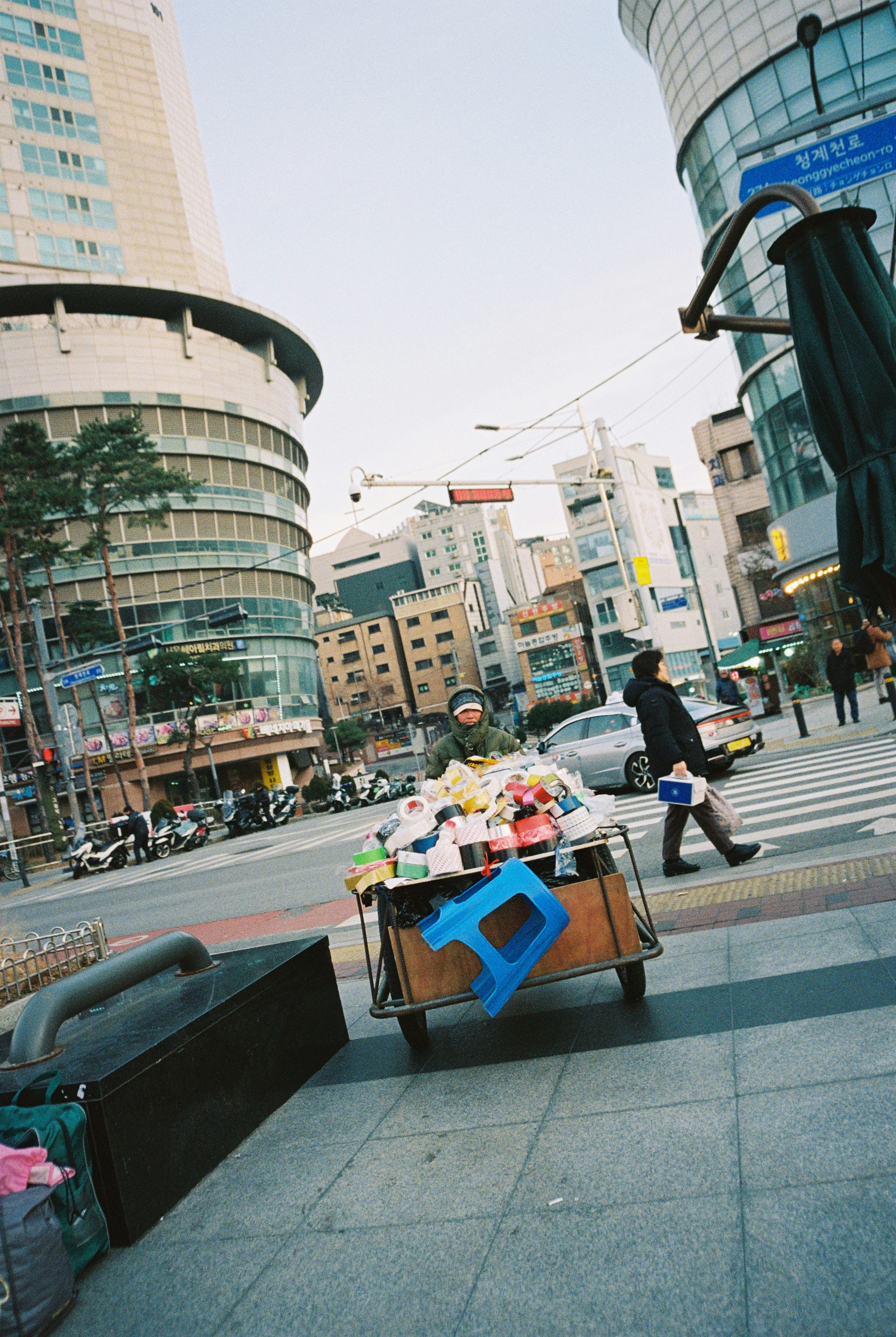 Colorful street cart laden with items sits at a busy urban crosswalk, with pedestrians and modern buildings in the background. A candid city scene focusing on everyday commerce.