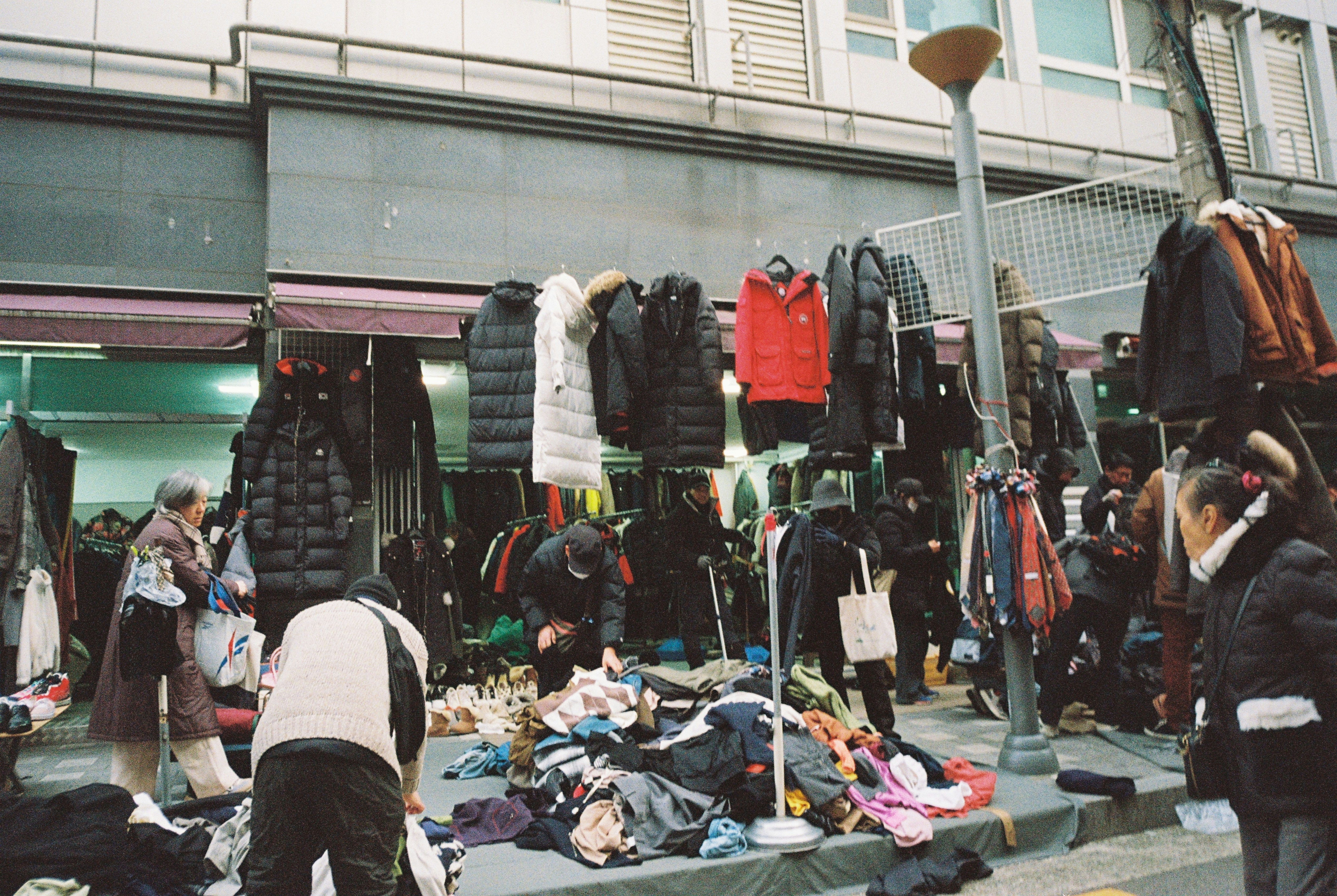 Vendors sort through clothing and jackets sprawled on a busy street stall at a market. A backdrop of storefronts and hanging garments frames a crowd of shoppers and workers in motion.