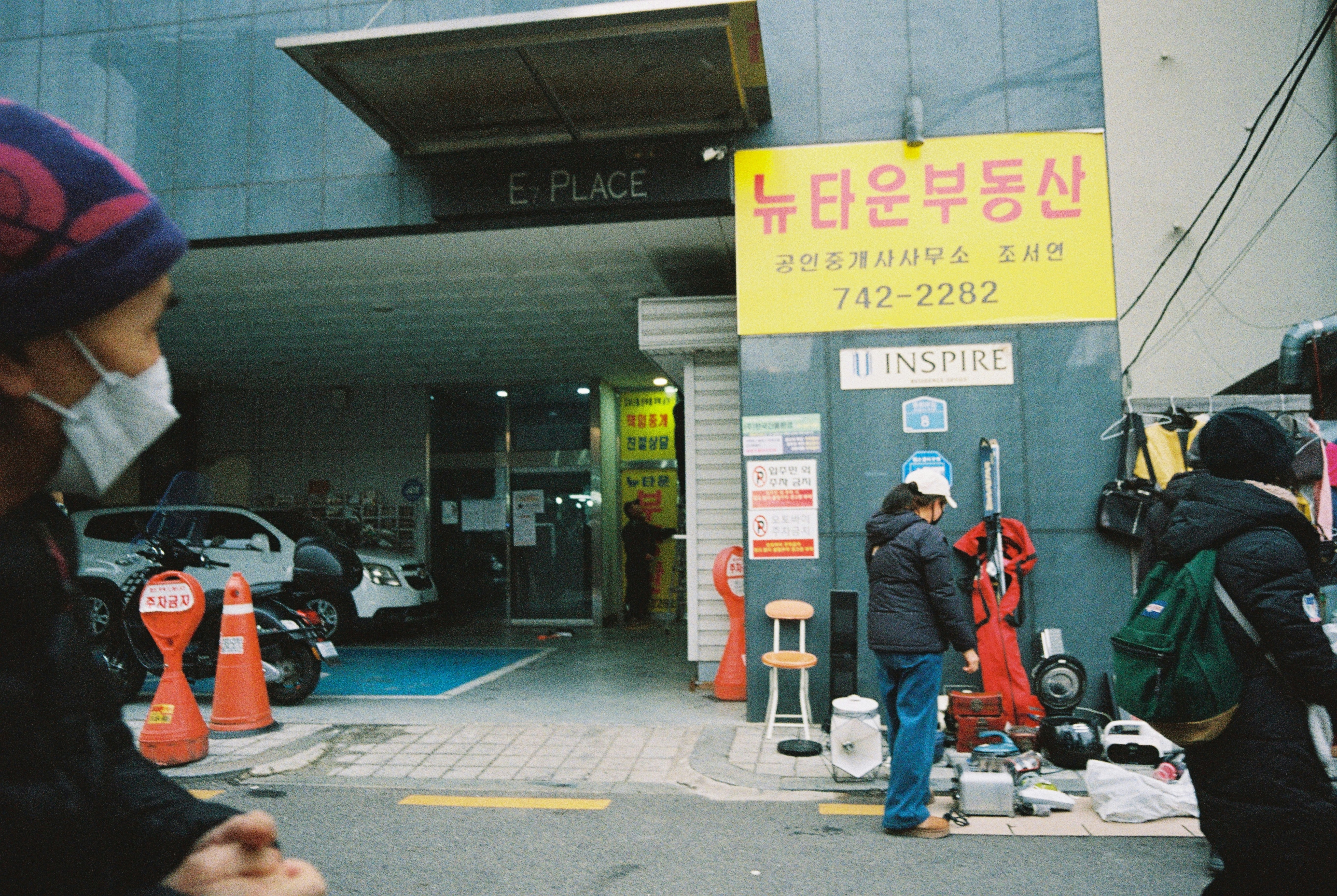 Busy storefront street scene with pedestrians and a yellow Korean sign; the composition centers on the doorway and nearby activity, including a man in a cap and mask and a vendor-like setup on the sidewalk.