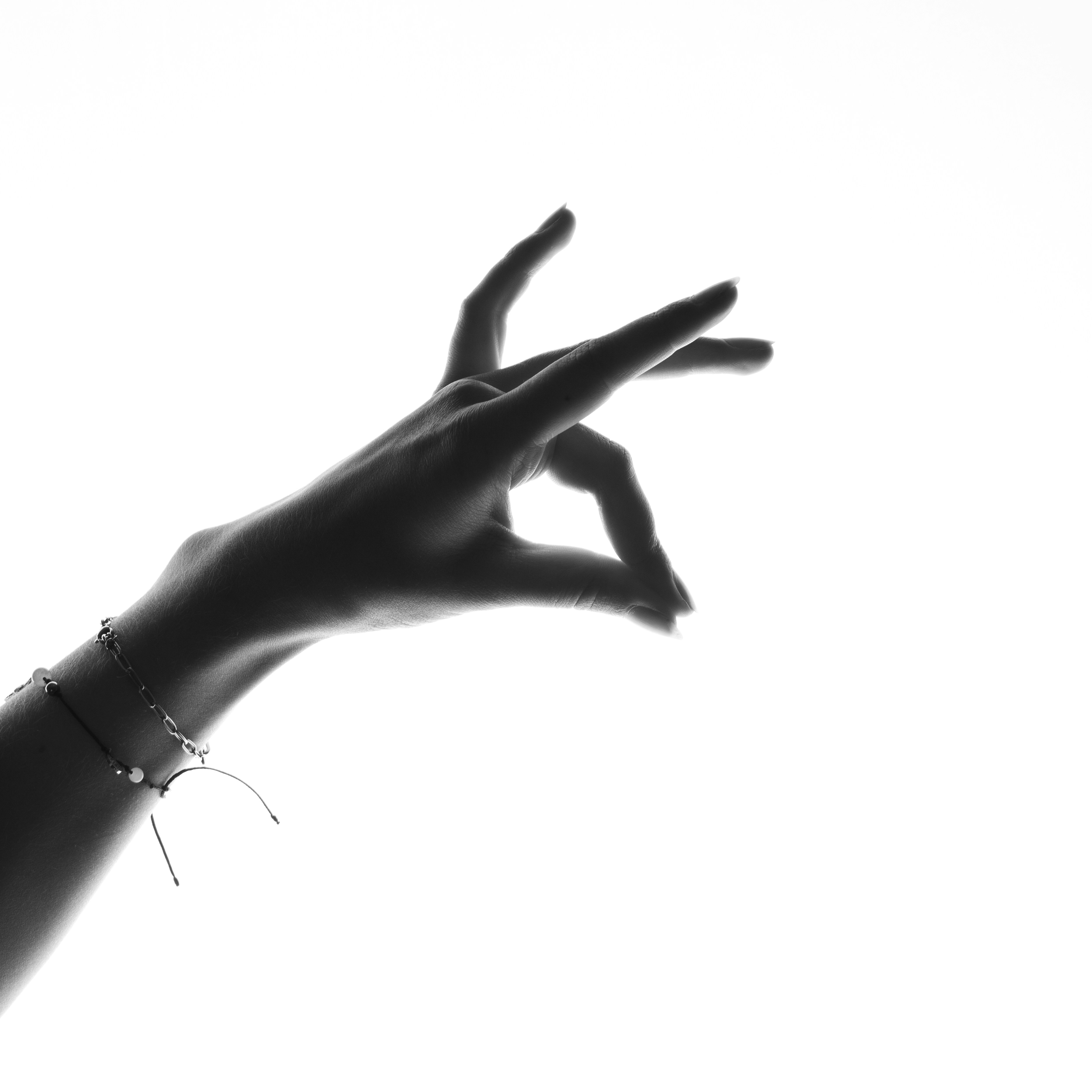 High-contrast silhouette of a left forearm and hand making a delicate gesture with a bracelet visible, set against a pure white background. Minimalist monochrome portrayal focusing on shape and negative space.