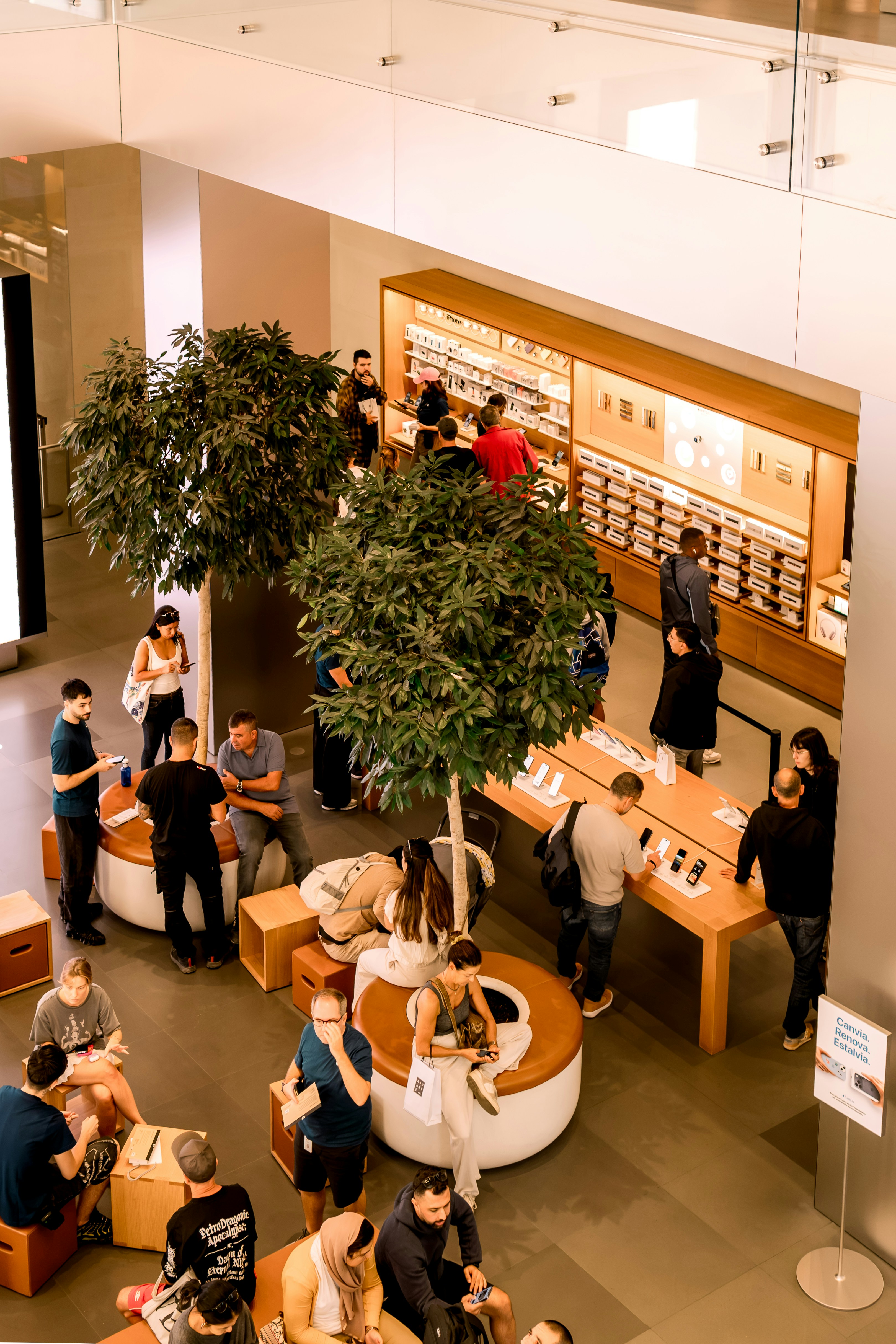 A crowded store interior with a central tree and circular seating around it, customers browsing and testing devices at wooden counters and shelves in a lit retail space.