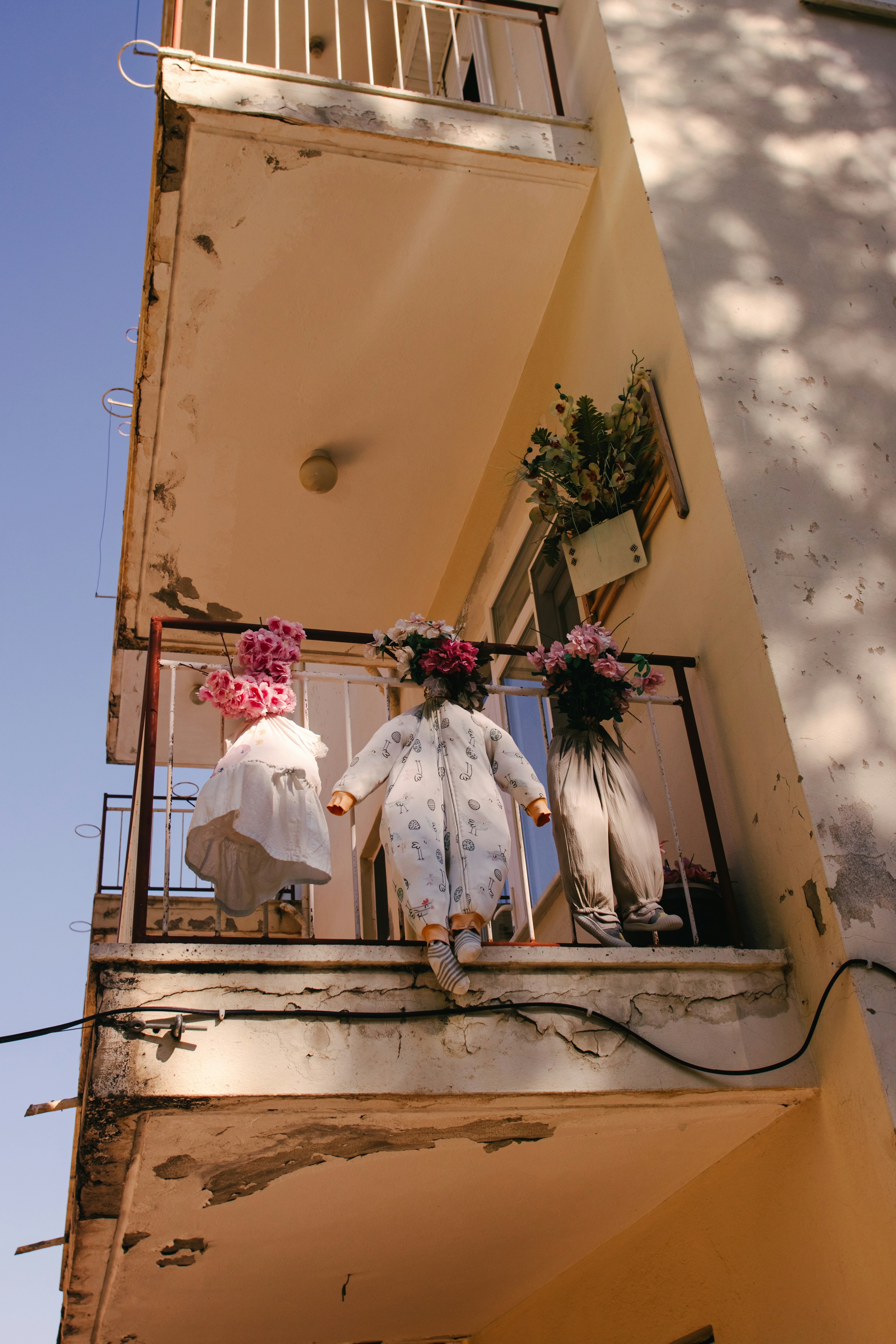 Three flower-adorned dolls stand on a sunlit balcony railing, their dresses flowing as they gaze outward against a pale building. A floral arrangement crowns each head, creating a whimsical still-life on an aging façade.
