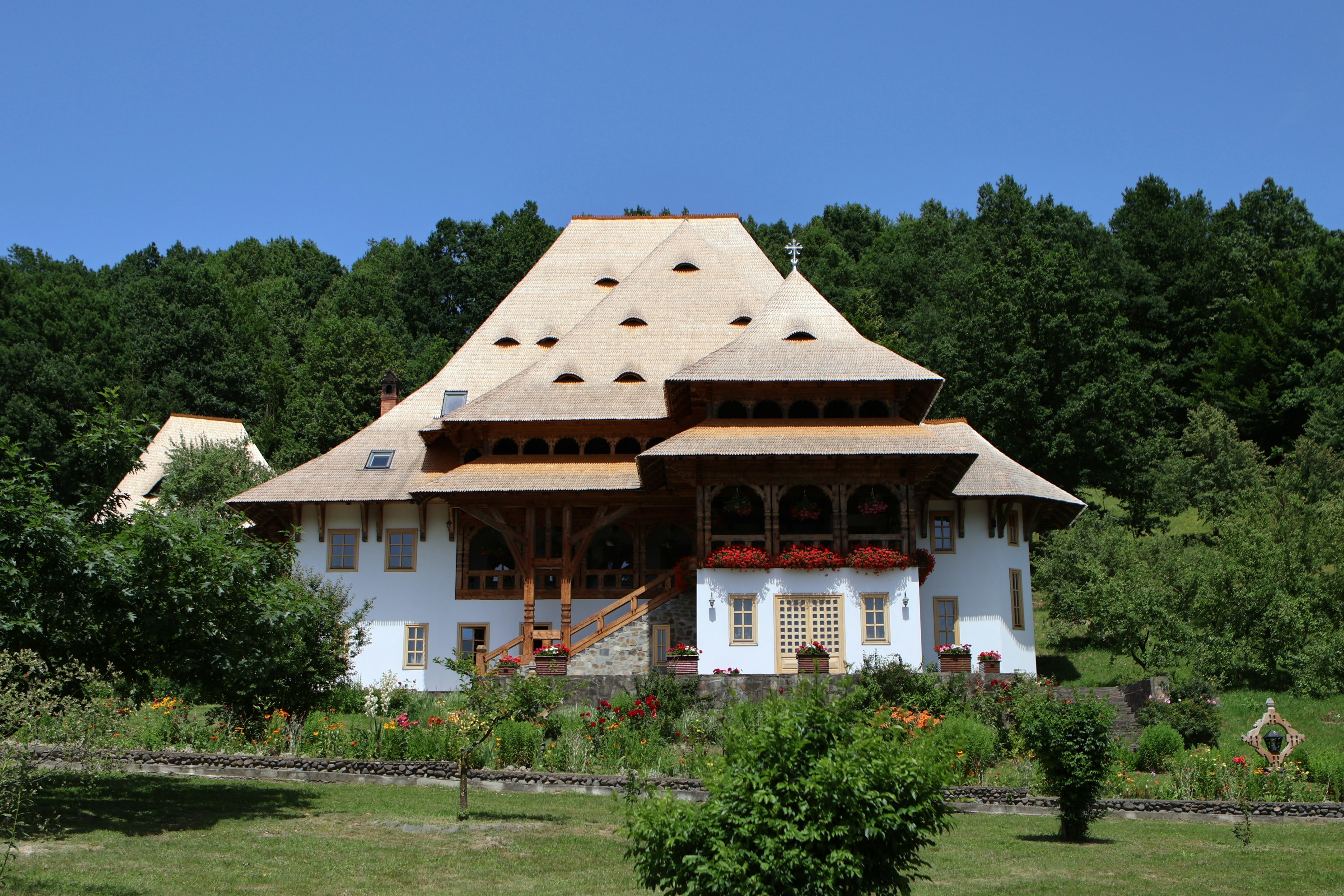 White wooden lodge with a steep thatched roof sits amidst green garden and forested hillside, showcasing traditional architecture with balcony and flower boxes.