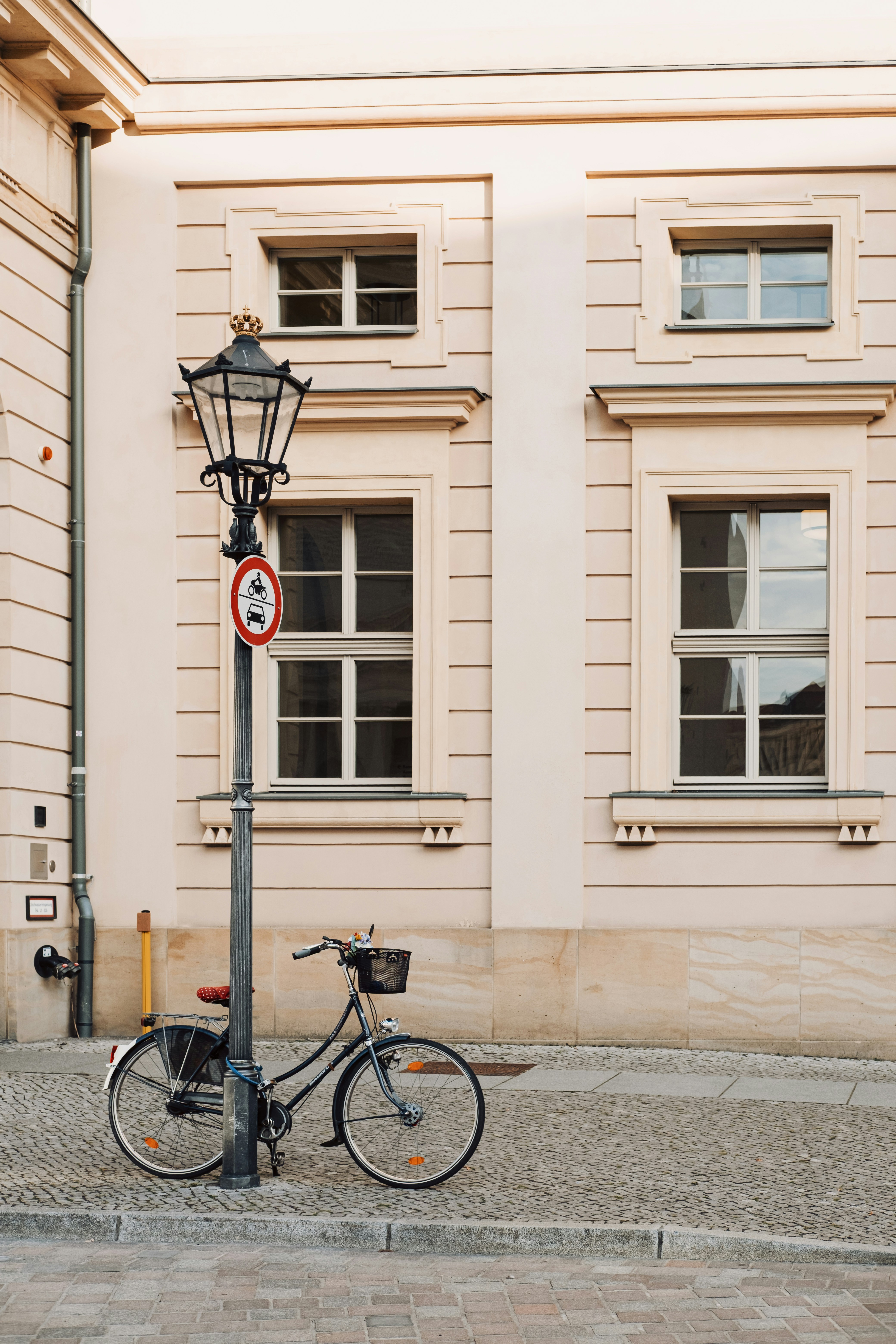 A lone bicycle locked to a vintage street lamp in front of a light-colored neoclassical building with tall windows. The scene captures urban still life with muted, warm tones.