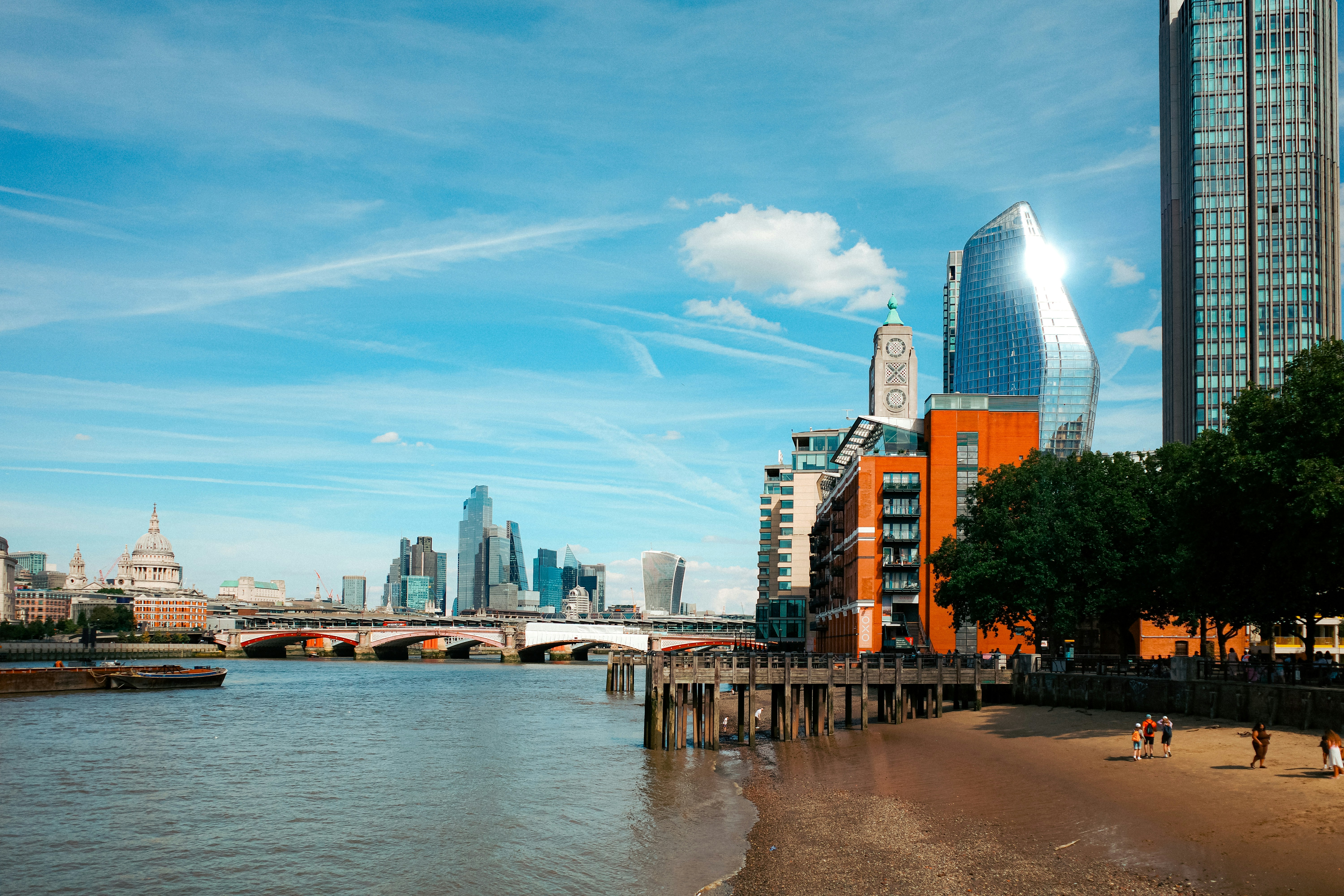 Wide riverside scene along a bustling urban waterfront with modern architecture and a clear blue sky. A sandy beach and pedestrians add human scale to the cityscape.