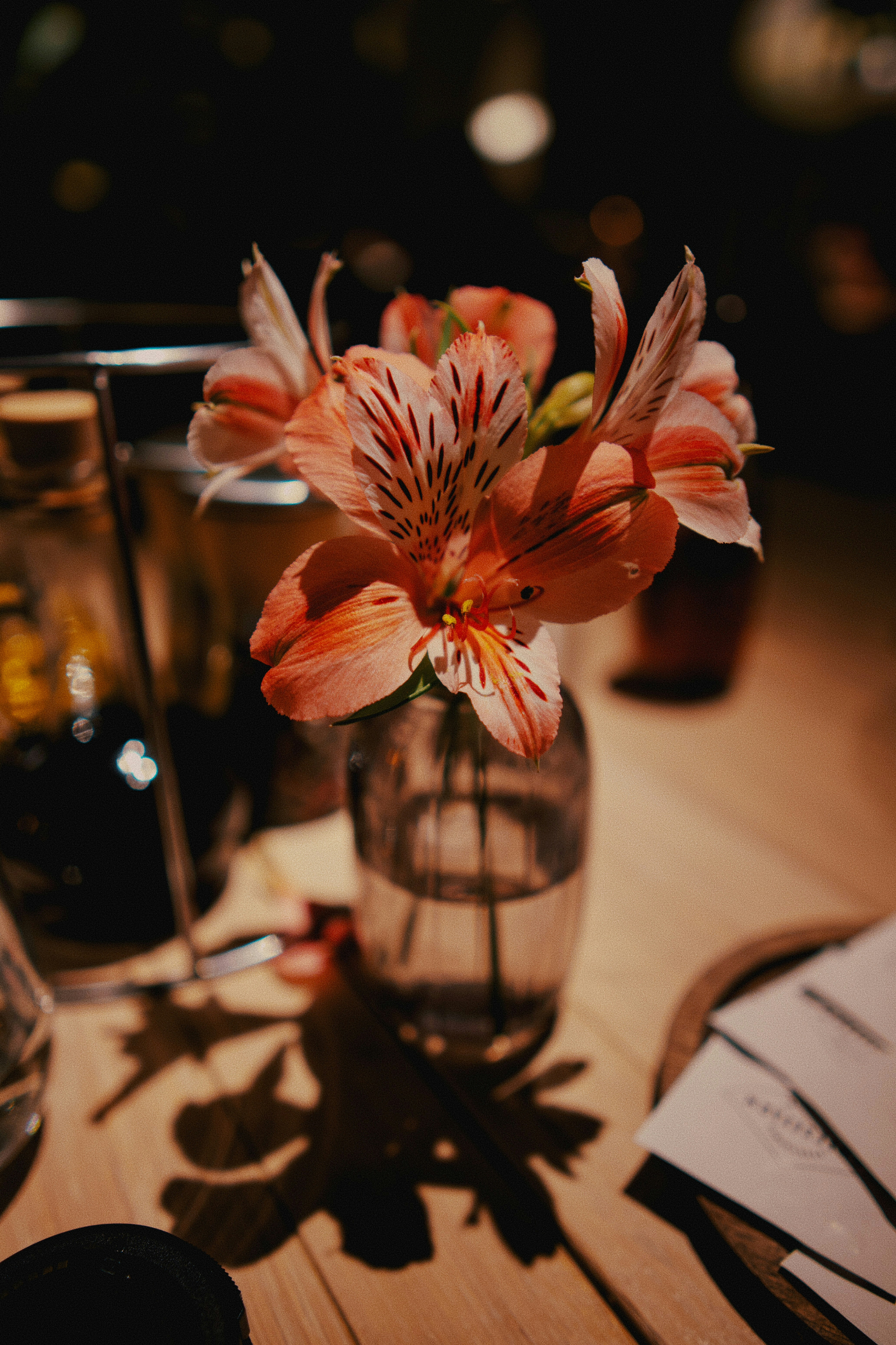 Softly lit flower arrangement in a glass vase on a wooden table; blurred bokeh lights in the background suggest a cozy dining setting.