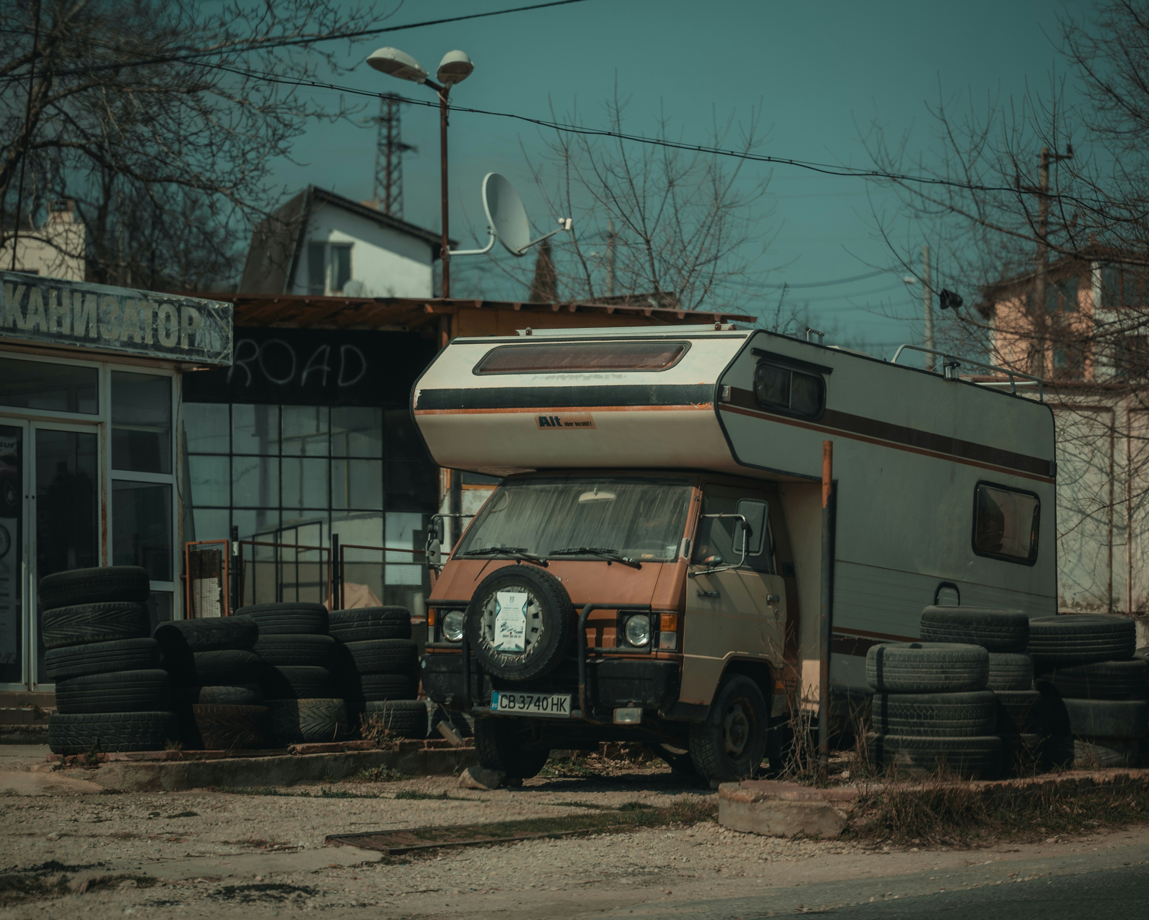Abandoned Road Warrior: A Camper’s Quiet Remnant Beside Stacked Tires
