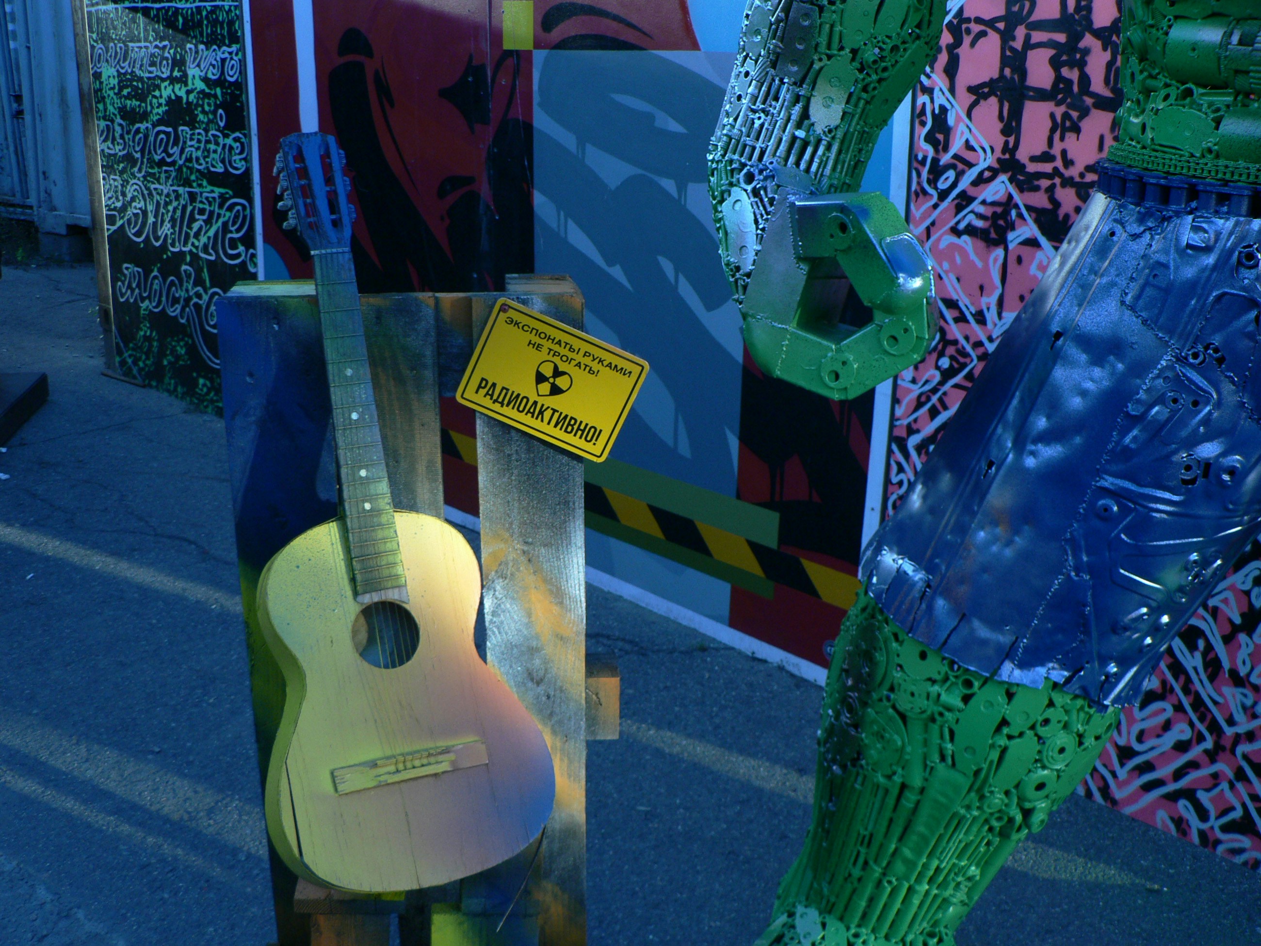 The image captures an urban art installation blending music and dystopian aesthetics. A wooden acoustic guitar with a warm, natural finish stands on a makeshift wooden pedestal, contrasting with the industrial elements around it. To the right, a large mechanical sculpture features vivid green modular limbs and a metallic torso, evoking a robotic or cyborg figure. A bright yellow warning sign with Cyrillic text (“Экспонаты руками не трогать! Радиоактивно!”) adds a layer of ironic hazard, while black-and-yellow caution tape reinforces the faux-danger theme.