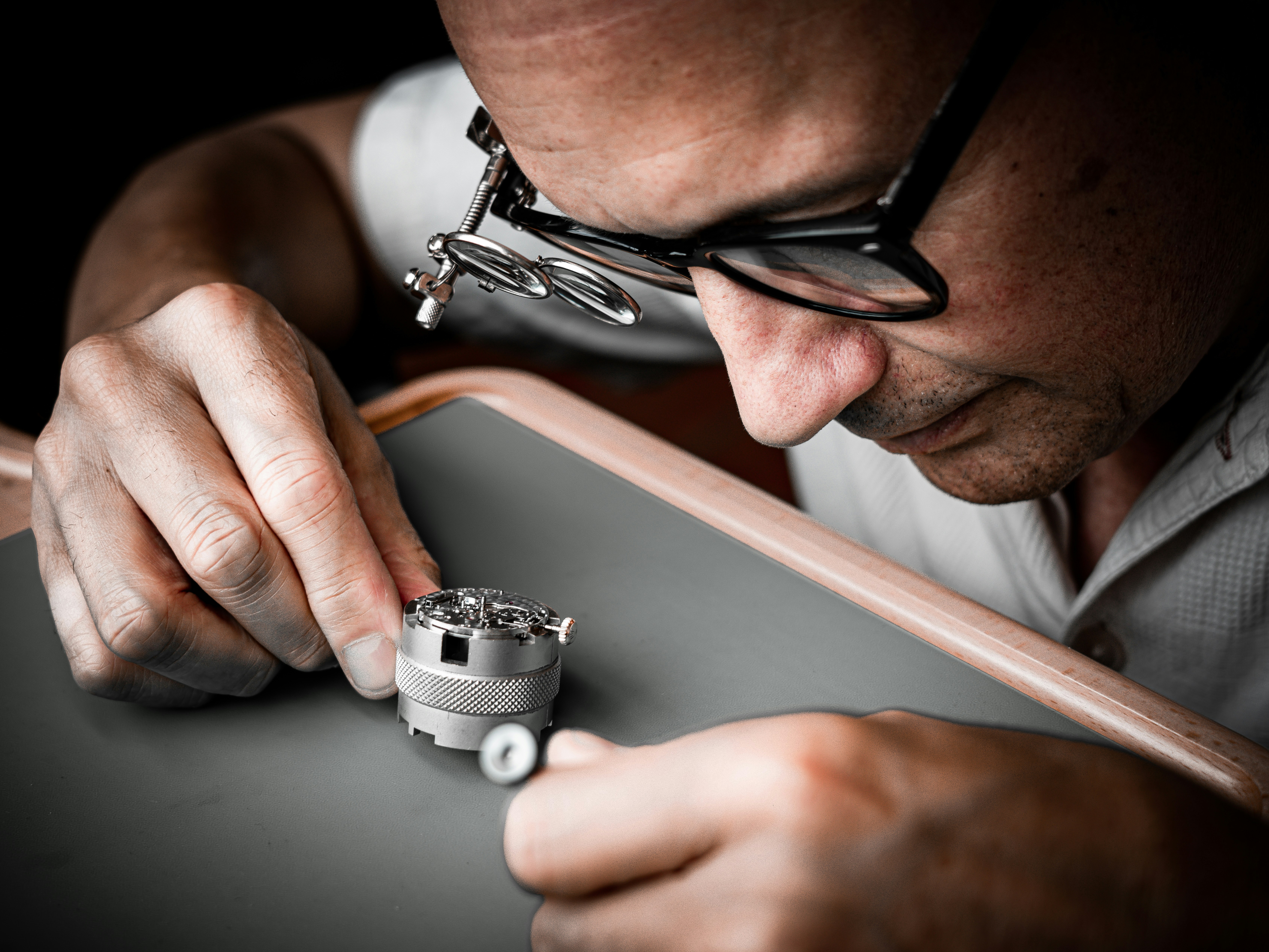 Close-up of a craftsman inspecting a precision component with magnifying glasses, the focus on hands and micro parts resting on a slate work surface. This is a meticulous, studio-shot setup.