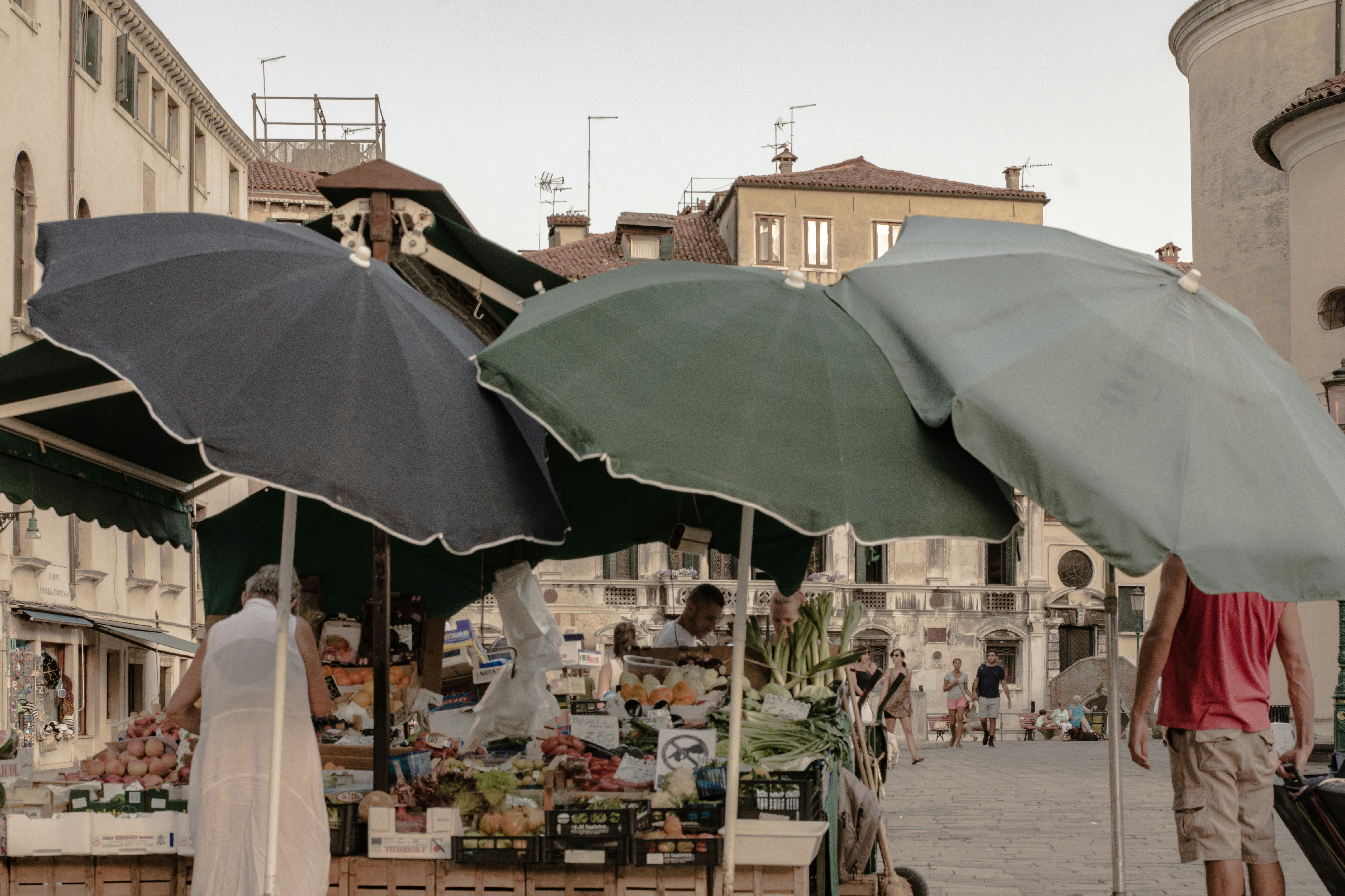 Bustling outdoor market under large umbrellas with vendors and shoppers, a stone-paved square and historic buildings in the background.