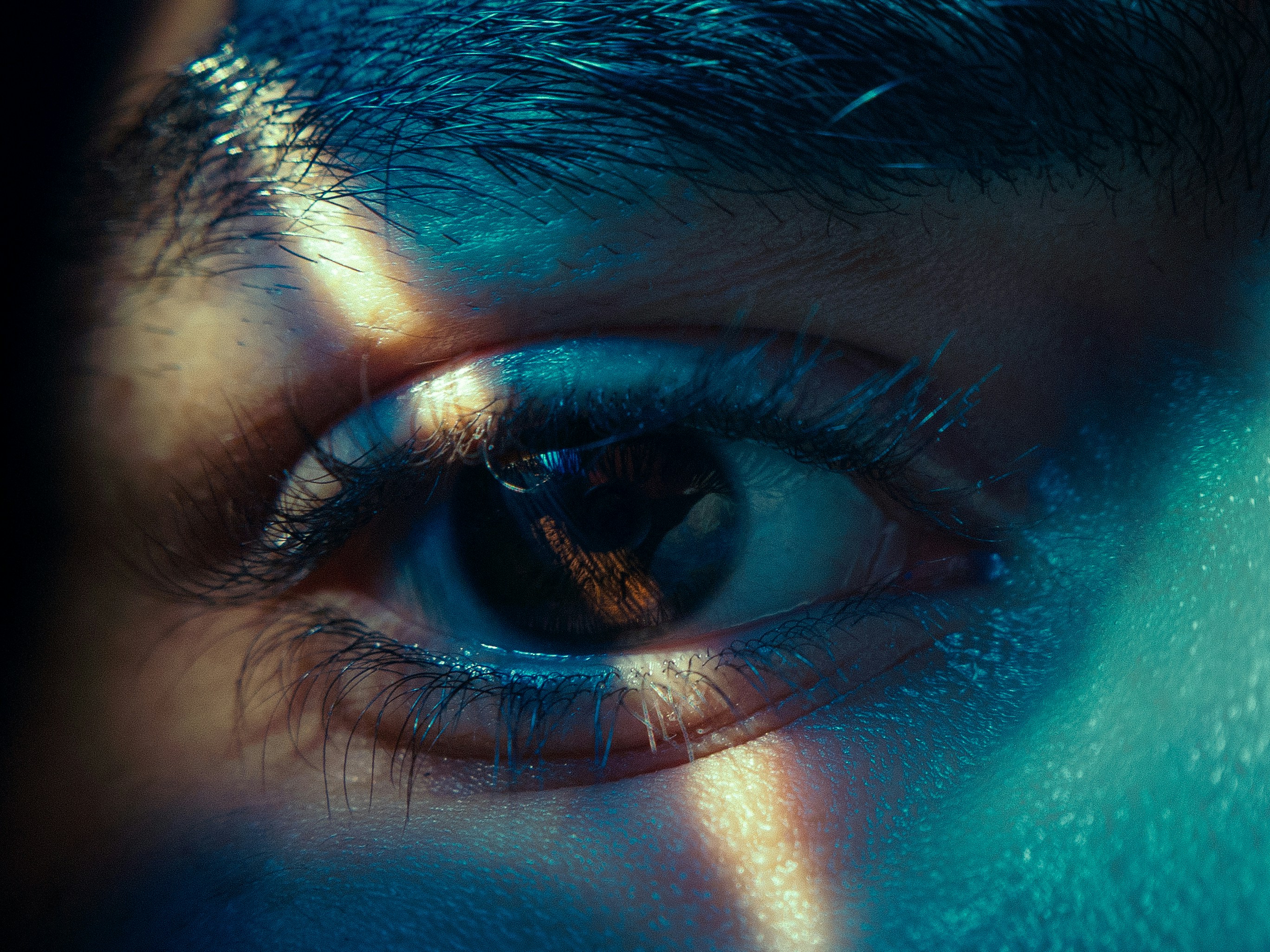 Close-up of a human eye with dramatic blue-green lighting and visible surrounding skin texture; the image emphasizes the iris and lashes with a moody, cinematic tone.