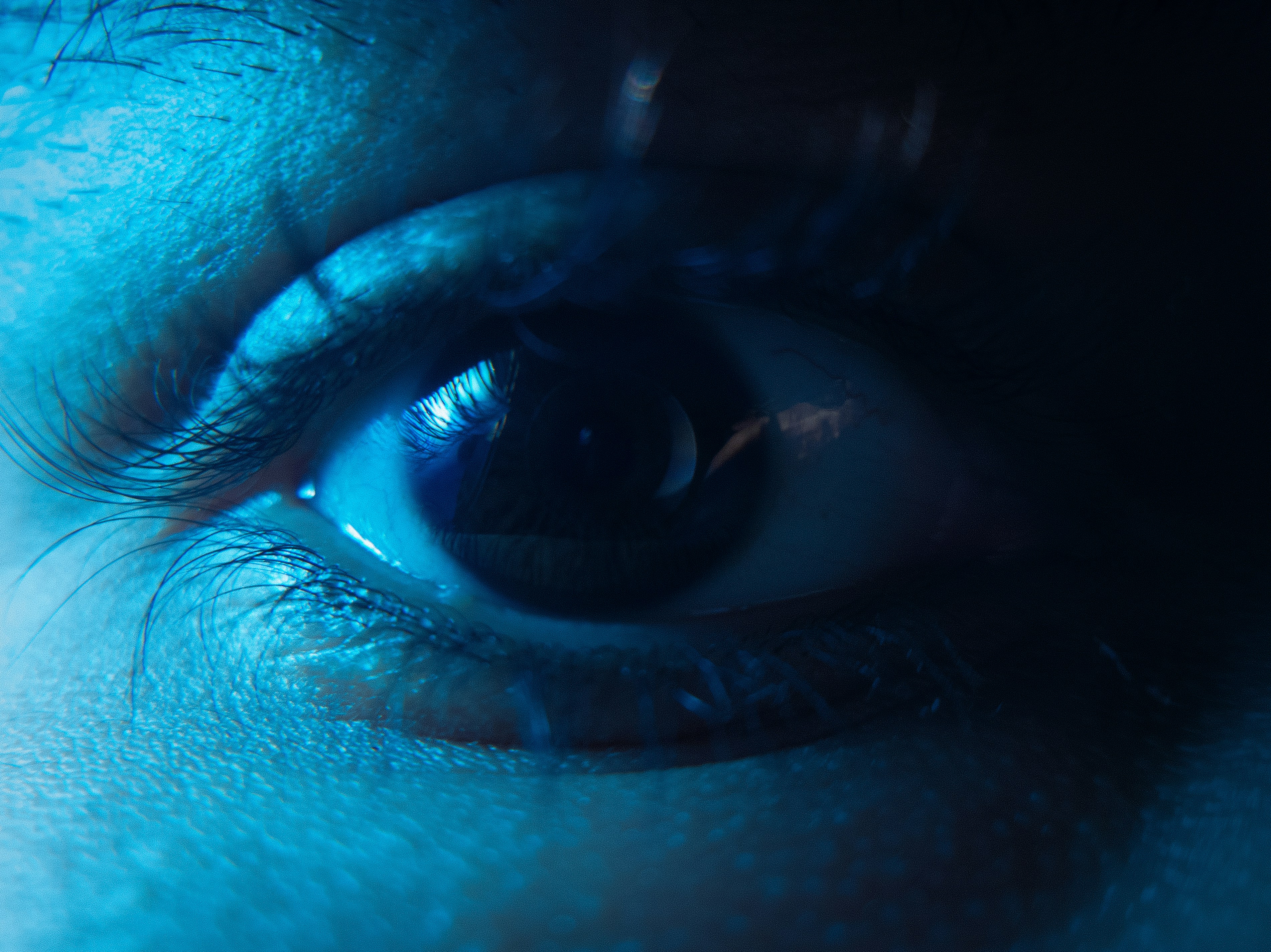 Close-up of a human eye bathed in blue neon light, revealing detailed iris and eyelashes; the image appears as a photographic capture rather than an illustration and emphasizes the eye as the focal point.