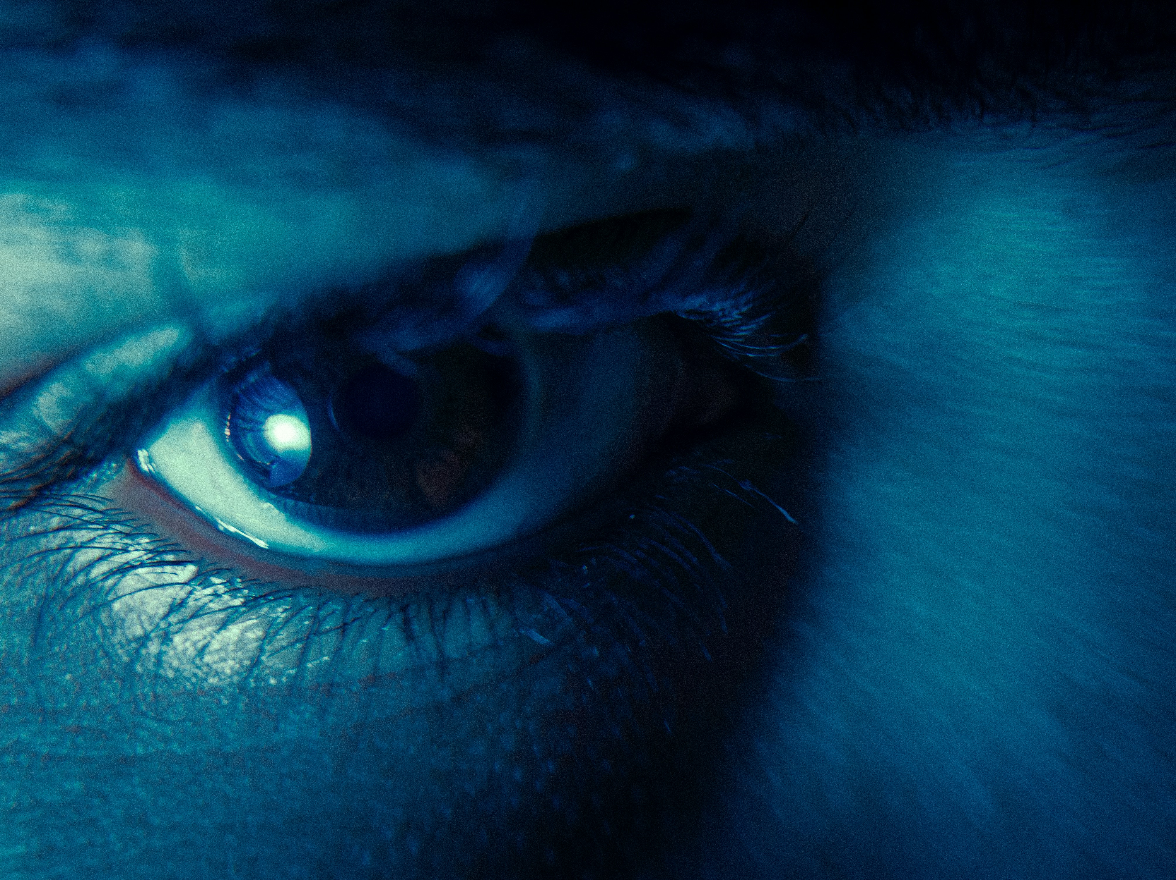 Intense close-up of an eye bathed in cool blue light, revealing detailed iris texture and lashes; likely a portrait with strong color grading in a controlled lighting setup.