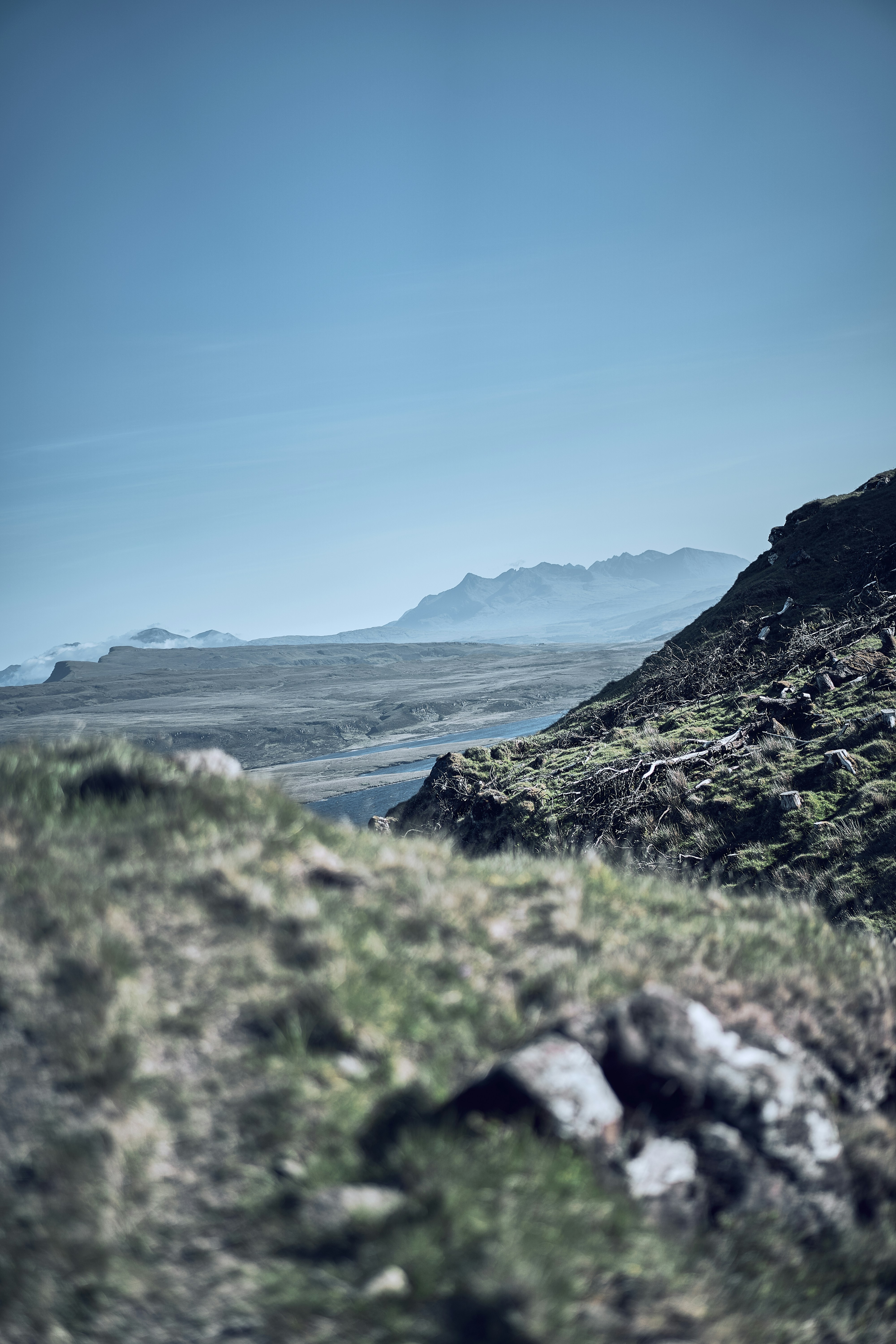 Rugged hillside foreground with a sloping rocky ridge leads to a distant volcanic plain and blue horizon.
