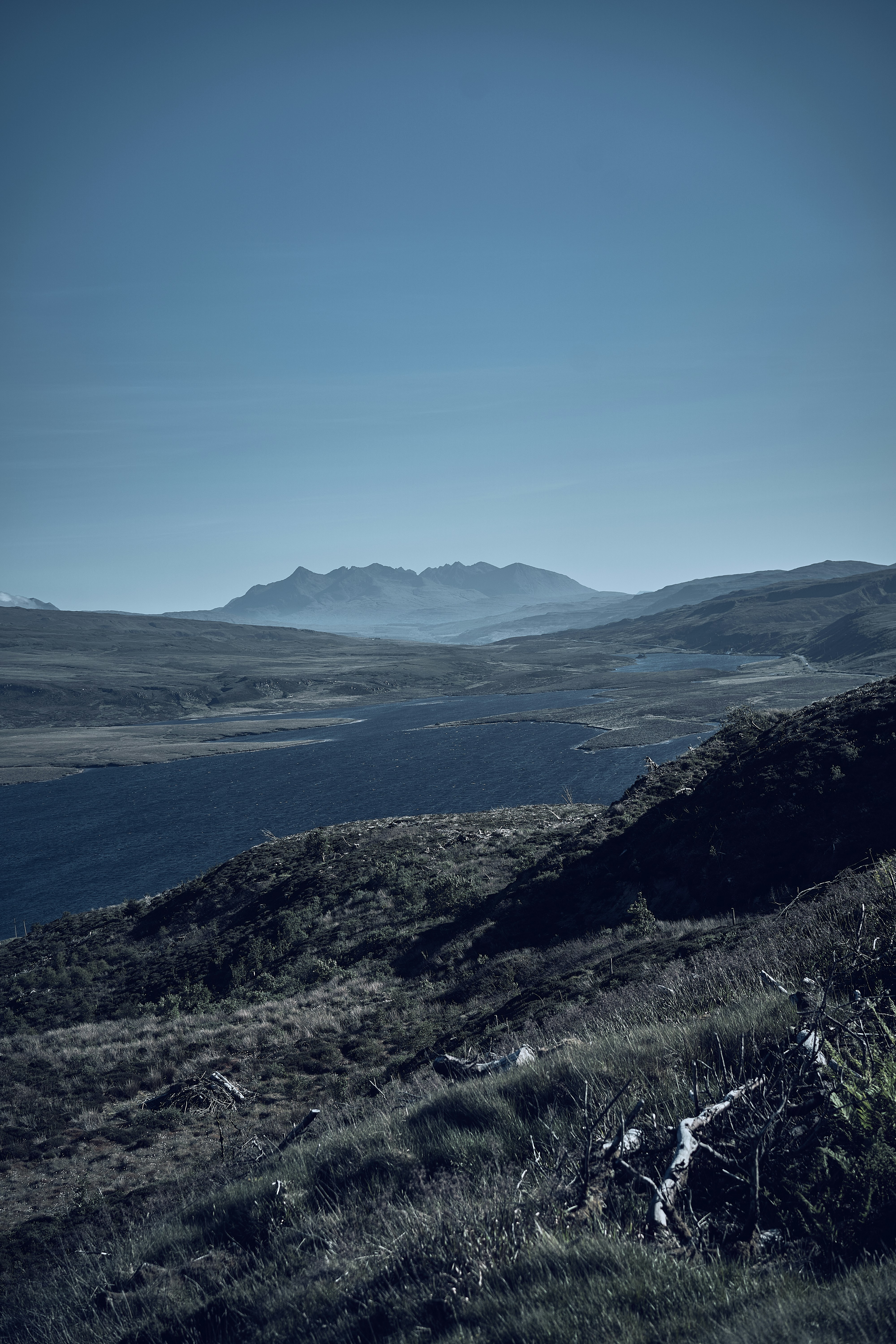 Open highland valley with distant mountain range under a pale blue sky; rocky foreground and rolling terrain lead the eye toward the horizon, conveying a sense of depth and solitude.