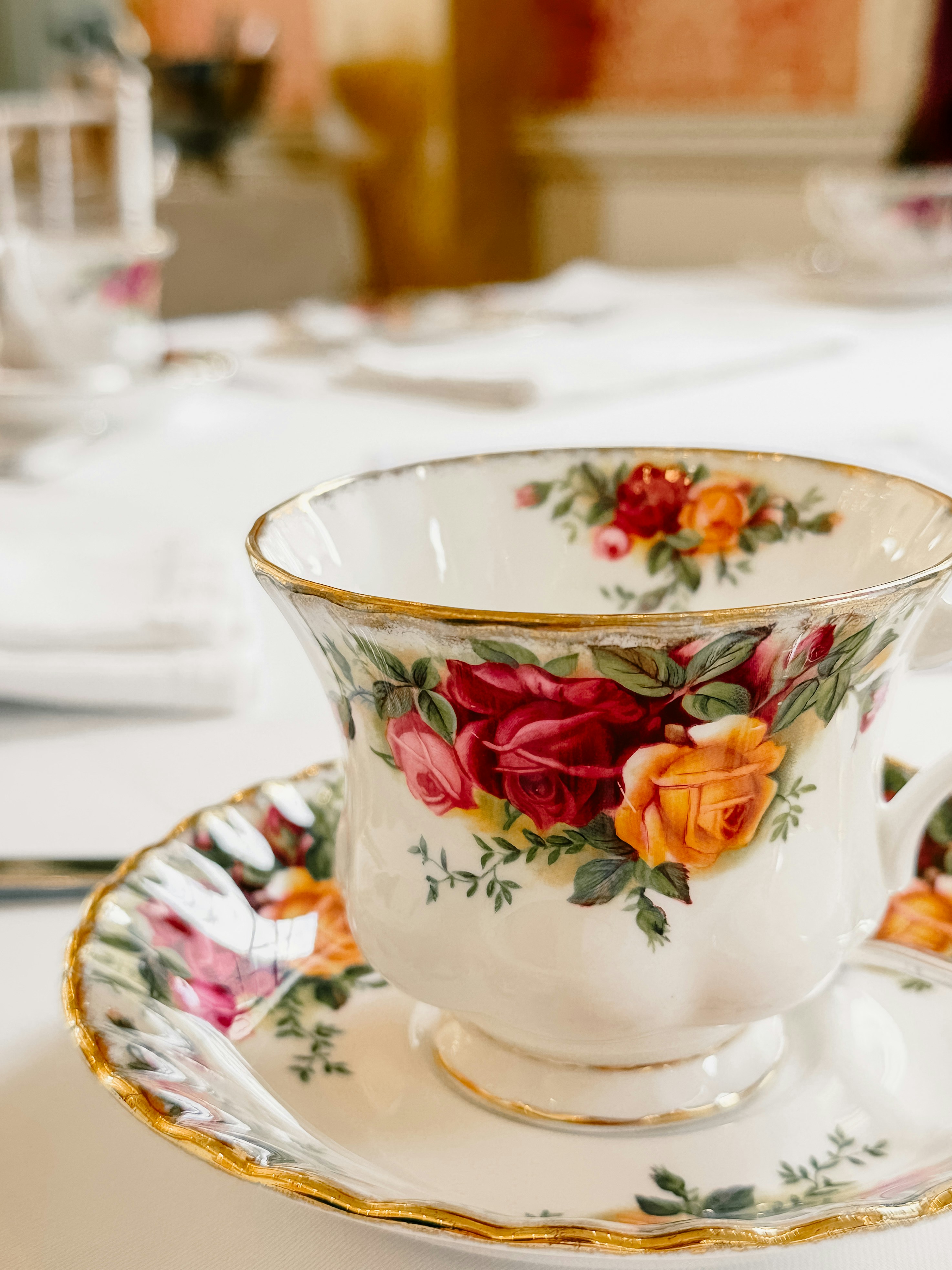 A fine porcelain teacup with rose motifs sits on a matching saucer, set on a white-draped table; the focus is on the cup’s detailed floral pattern and gold trim.