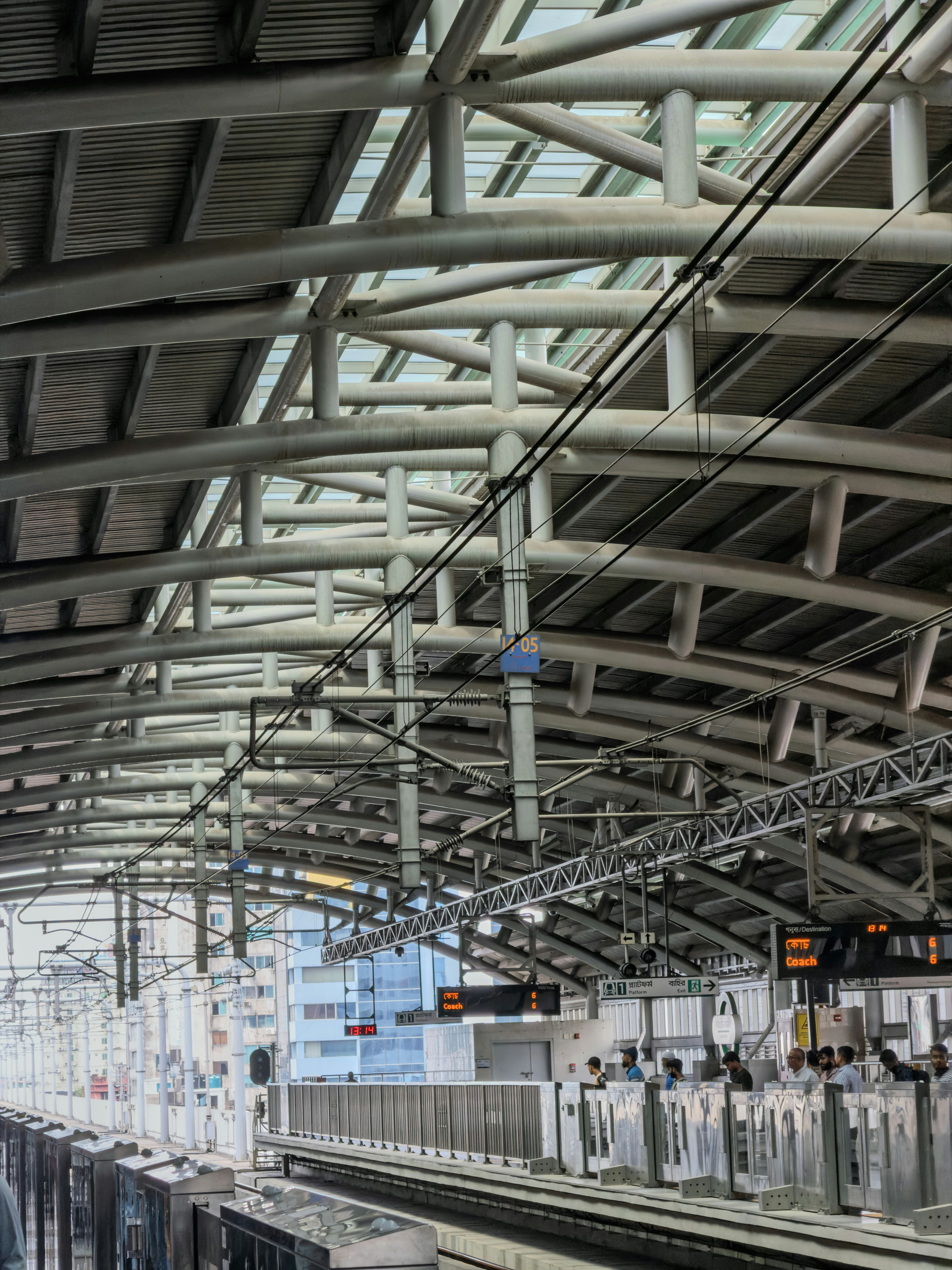 Platform with glass barriers beneath a network of steel arches and overhead wires at a city train station.