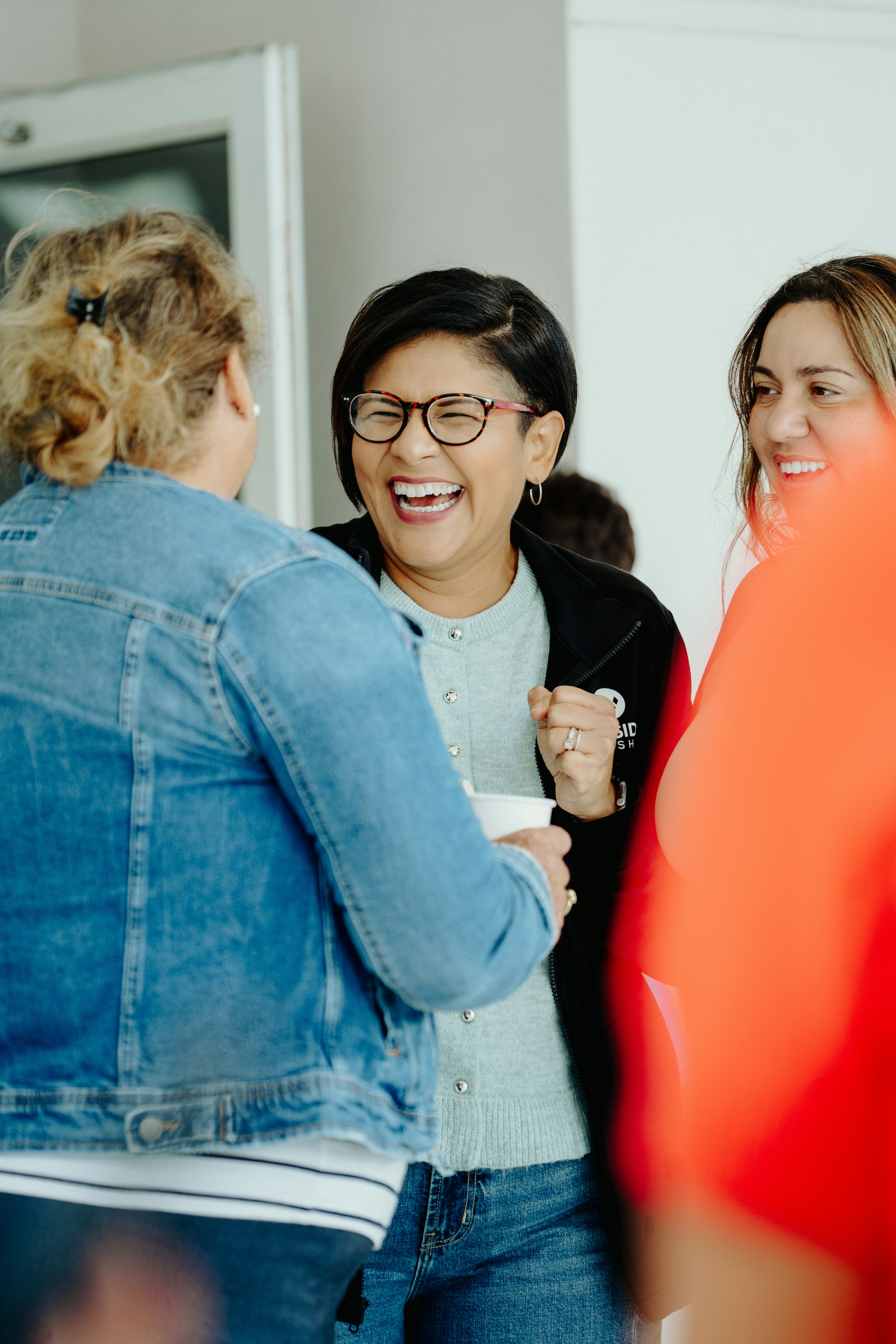 Three women in conversation laugh together at a casual indoor event; one wears a denim jacket, another with glasses and a light cardigan, a blurred red shape in foreground hints at movement.