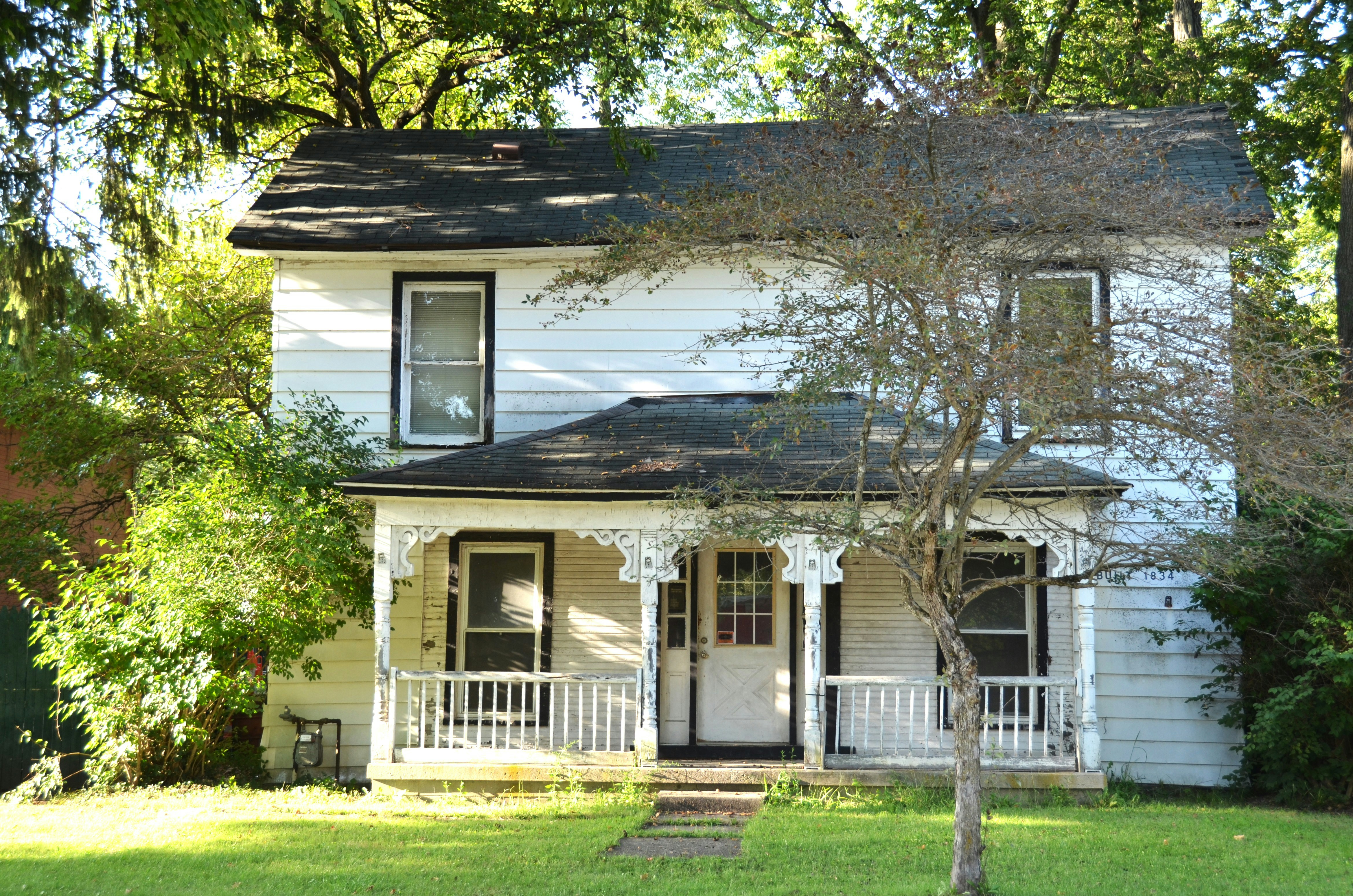 Two-story white wooden house with a covered front porch and decorative trim, set on a grassy yard with a bare tree in the foreground.