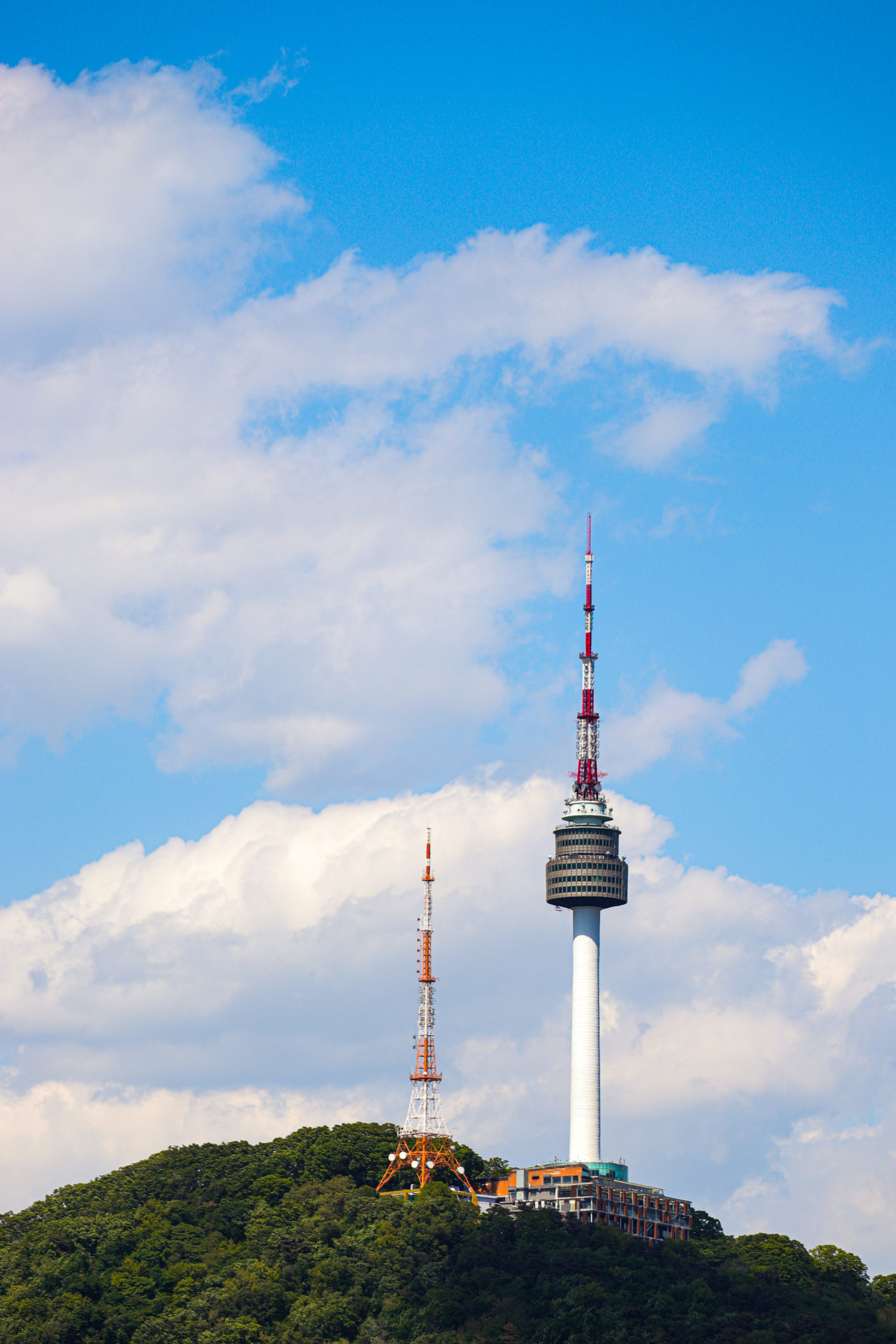 A tall communications antenna rises above a grassy hill against a bright blue sky with fluffy clouds; the structure sits near a cluster of buildings, creating a prominent skyline feature. This photograph captures the tower as the focal point of the scene.