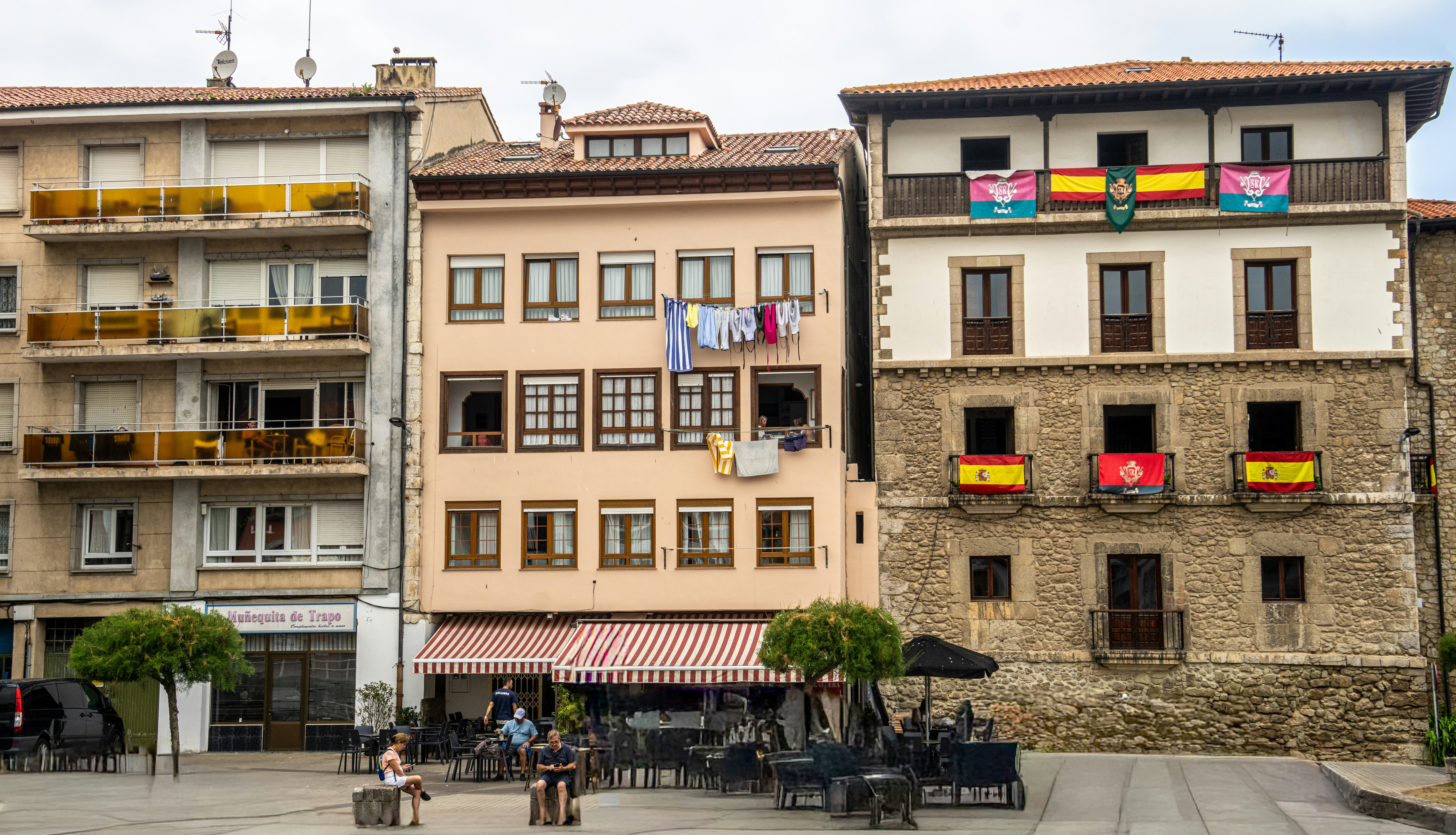 Colorful row of buildings with balconies and flags lining a sunny square; outdoor café tables and people lounging near the storefronts. A mural-adorned top-floor adds character to the scene.