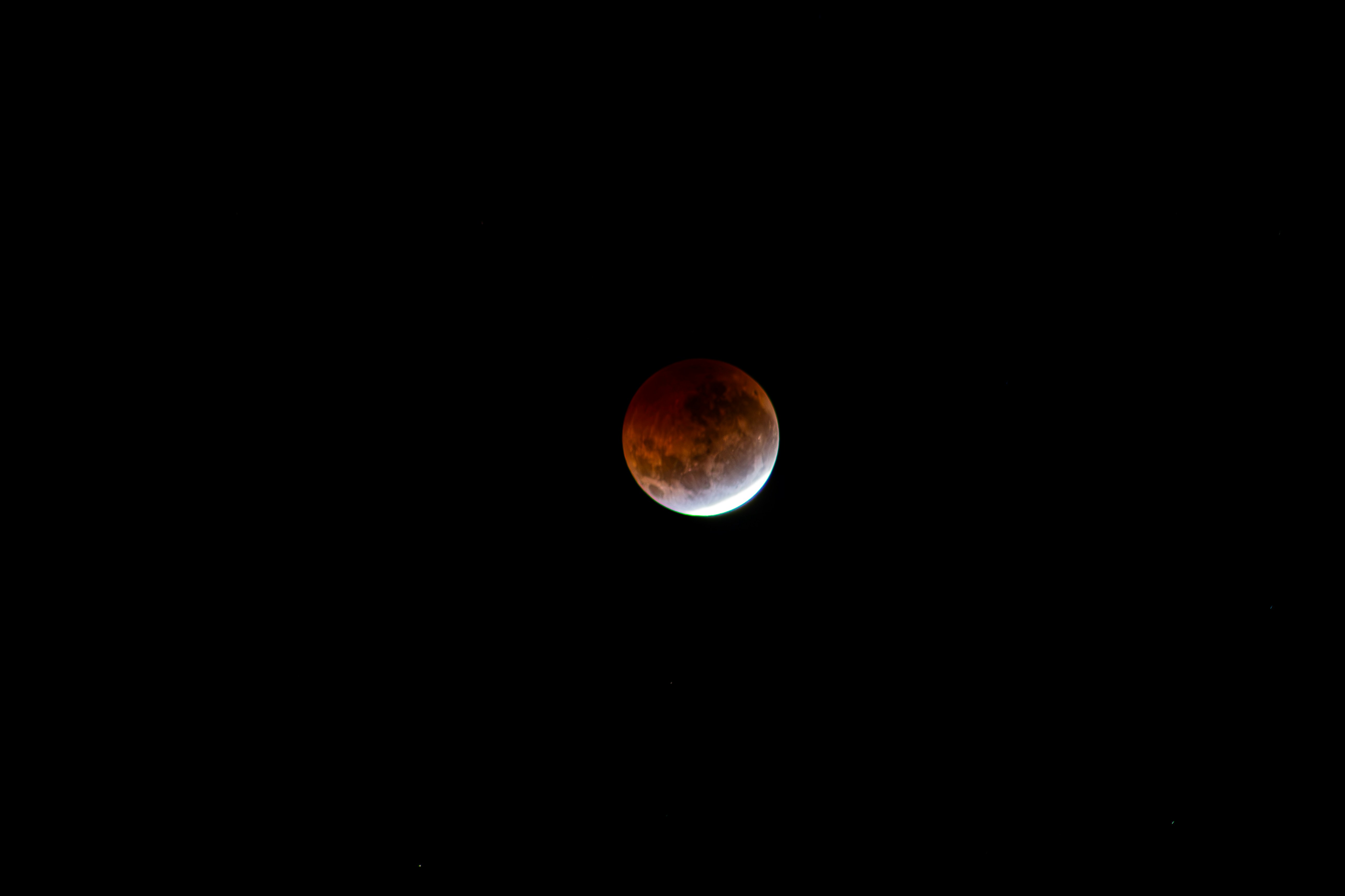 The eclipsed Moon glows rust-red over São Paulo as a bright crescent of unshadowed surface returns along its lower edge, marking the end of totality. The empty black sky isolates the disc completely, with only a few faint stars registering in the frame.