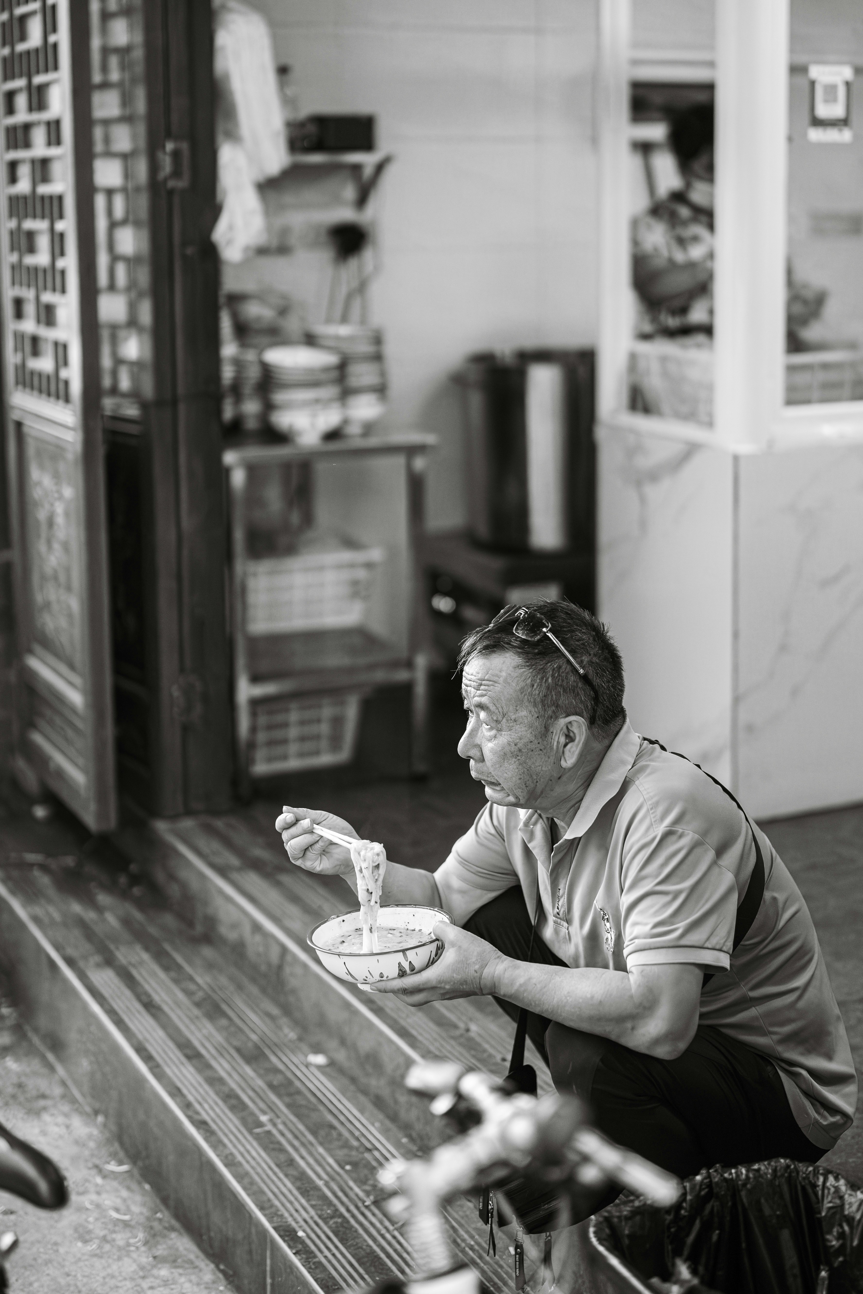 A man crouching on a step eating noodles from a bowl with chopsticks