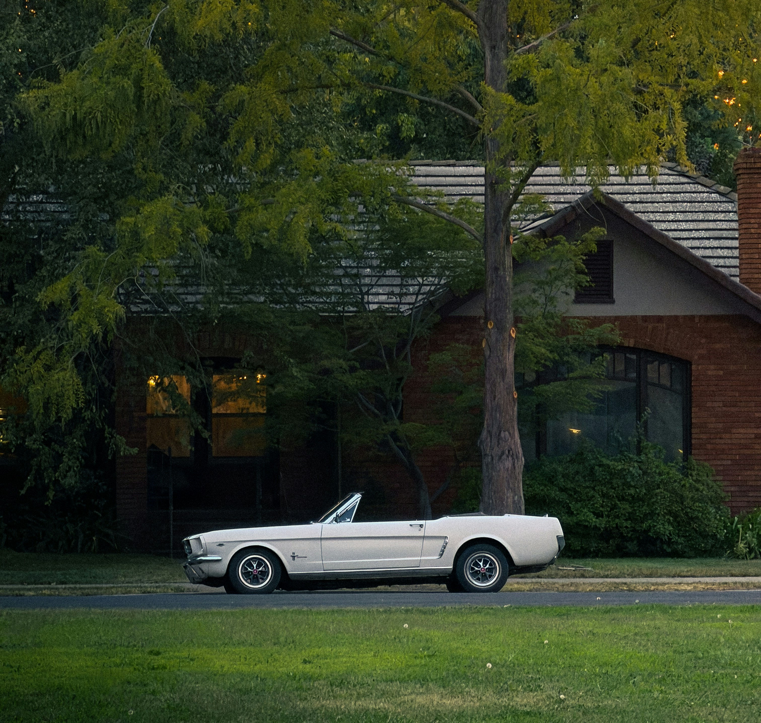 White vintage convertible parked along a suburban street with a brick house and trees in the background.