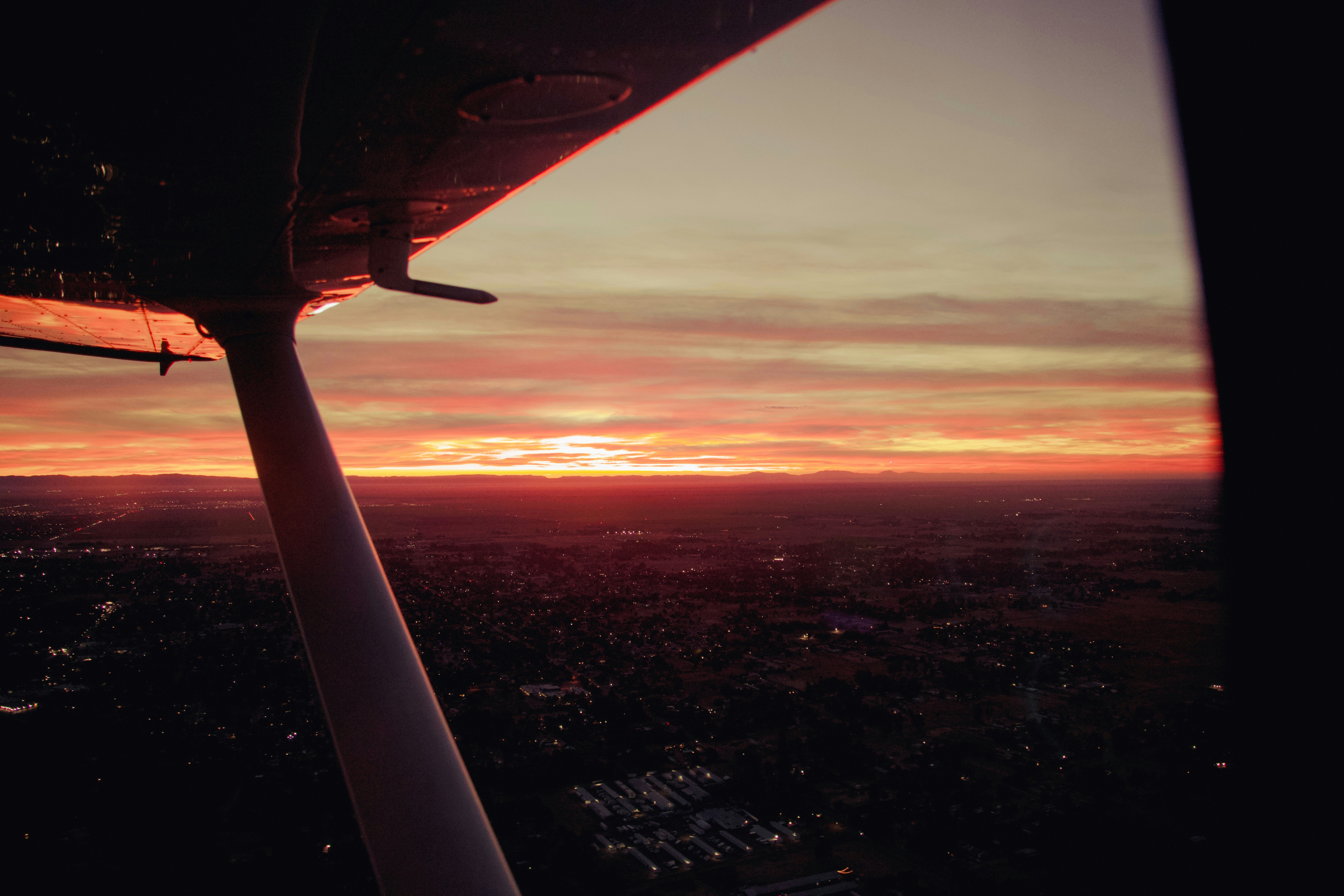 Dusk light washes the horizon with warm orange tones as airplane window framing and wing strut silhouette the scene; deep city lights speckle the landscape below. This is a photographic frame from a cockpit window, capturing the aircraft’s interior framing and the outside sunset.