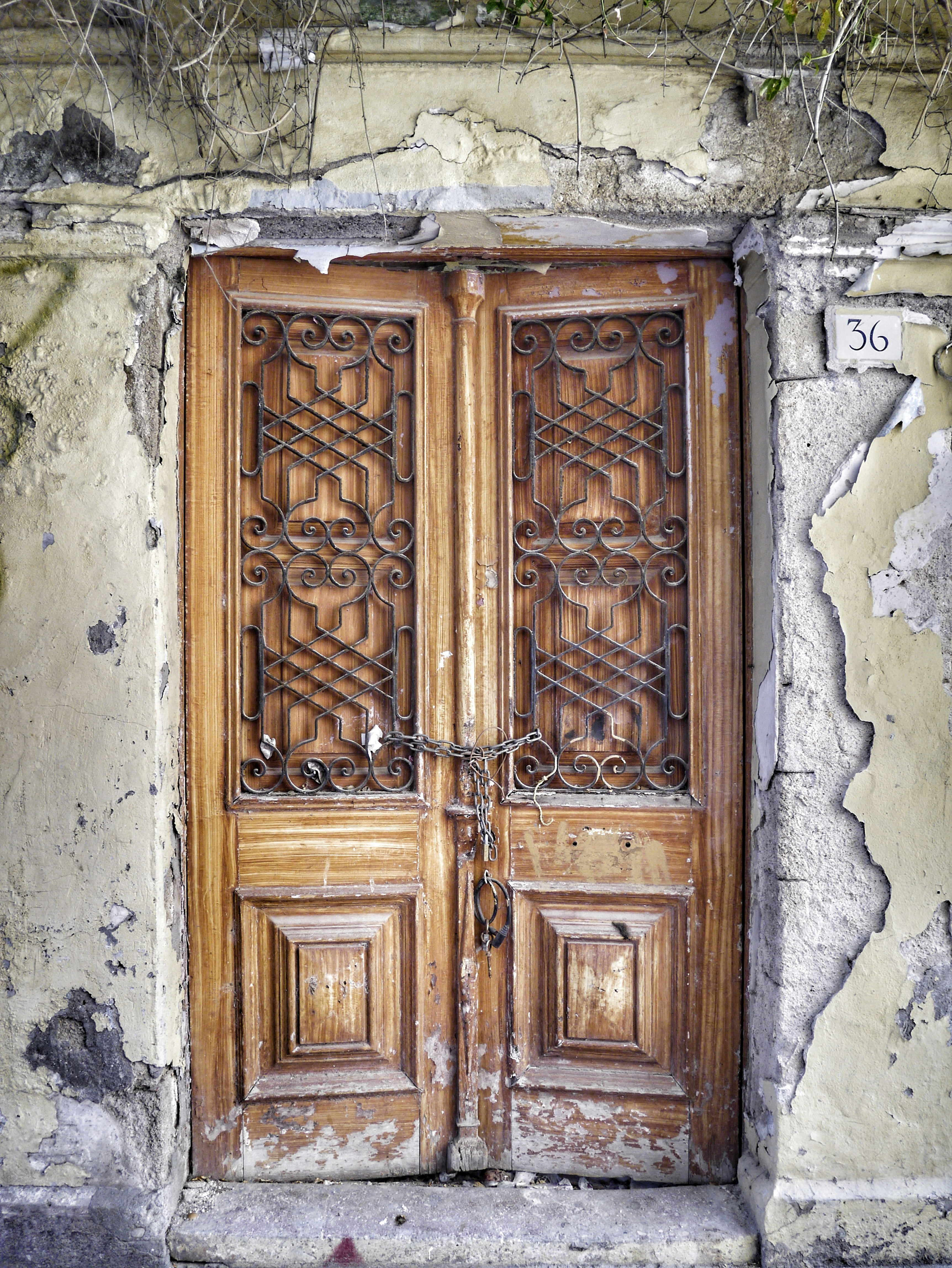Sun-bleached wooden double door with ornate iron grille panels, chained and weathered by years of exposure; crumbling plaster and faded numbers frame a neglected entryway.