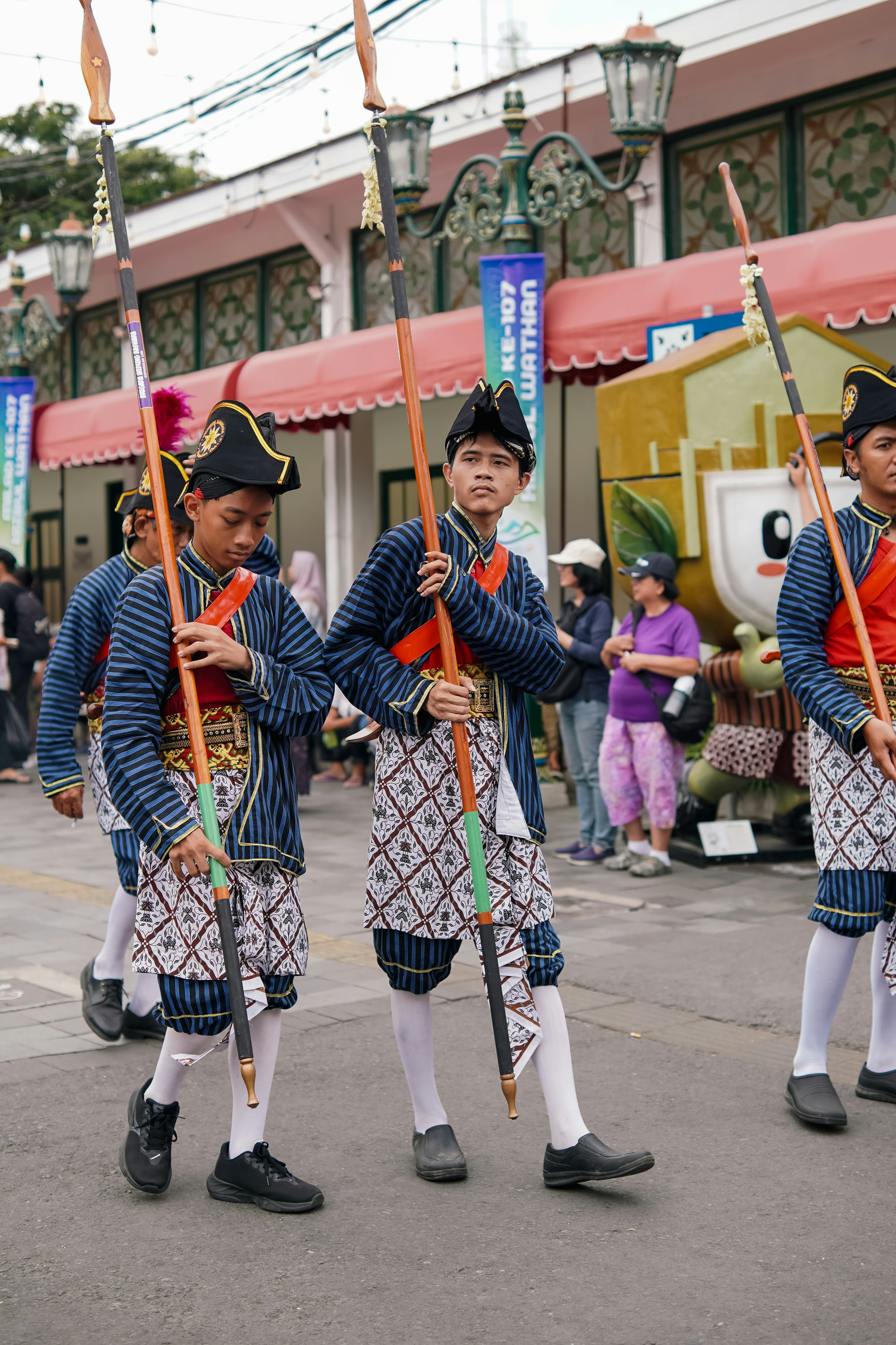 Spear-bearing performers in traditional attire march along a street during a cultural parade, with spectators and storefronts in the background. The scene captures coordinated movement and festive clothing.