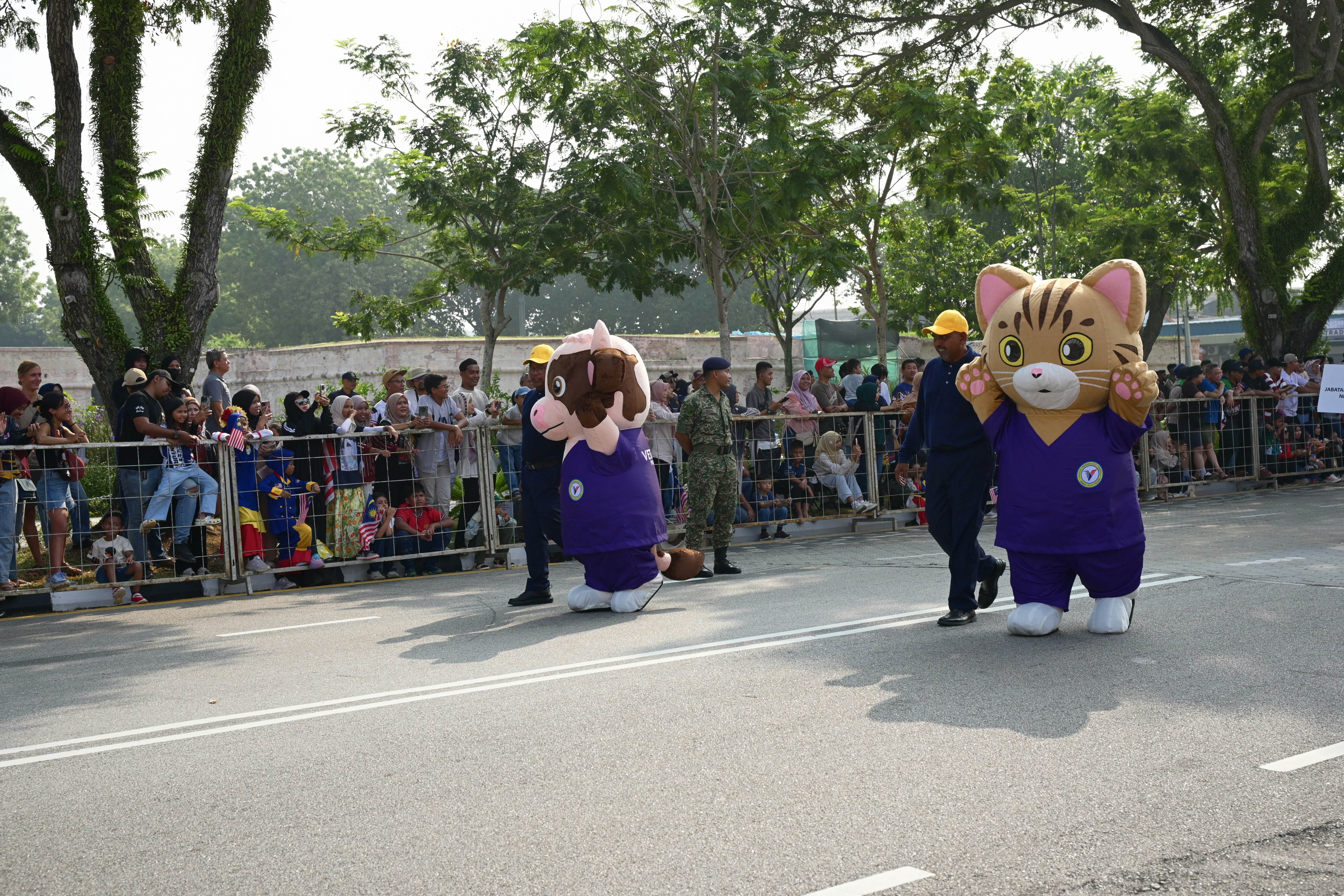 A street parade with costumed mascots in purple outfits walks along a closed road, spectators behind barriers, and a few uniformed staff nearby. Colorful characters dominate the scene in daylight.