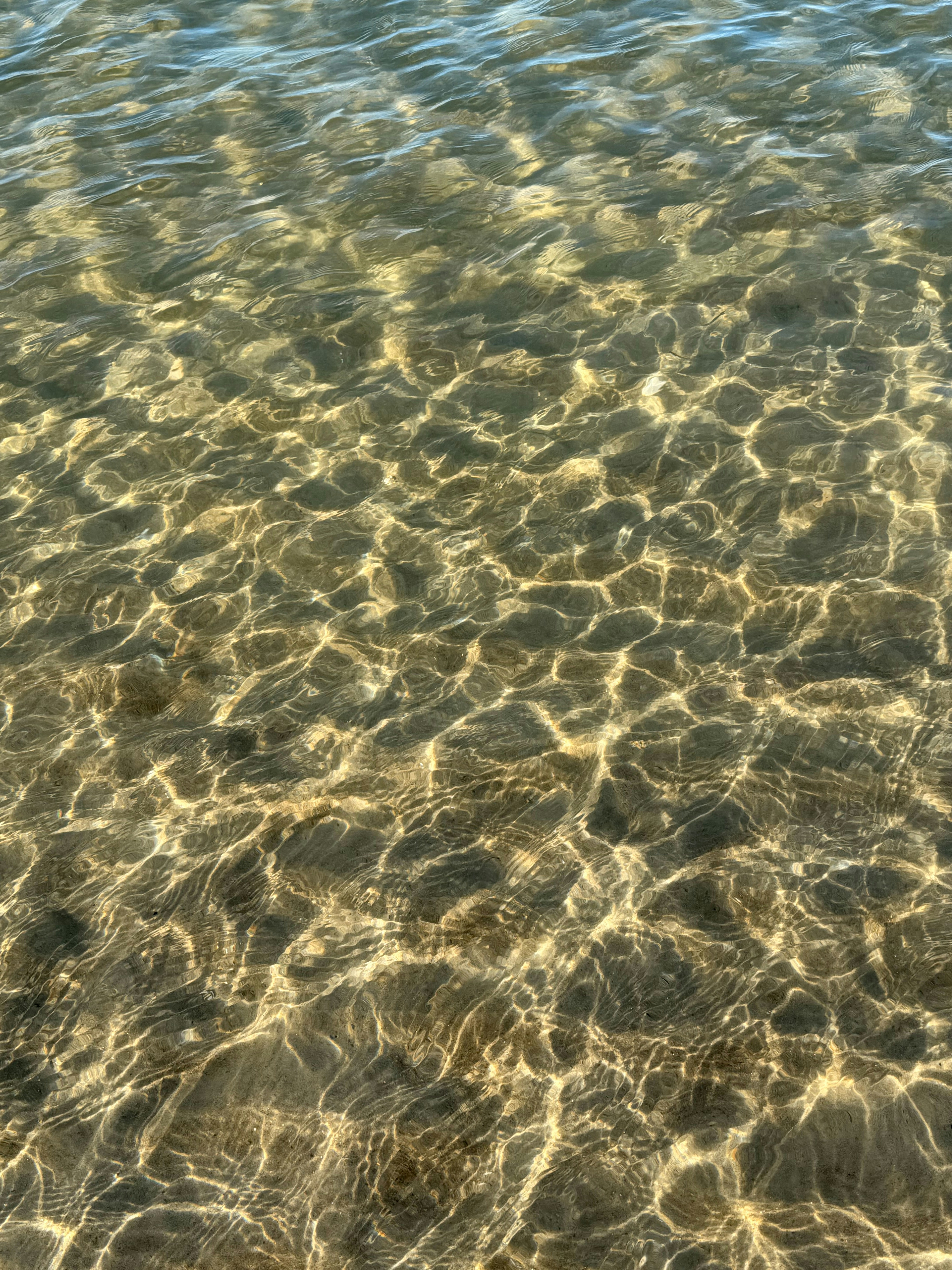 Sunlit ripples over sandy shallow water create a pattern of gold and shadow in calm surf. A close-water abstract highlighting texture and light.