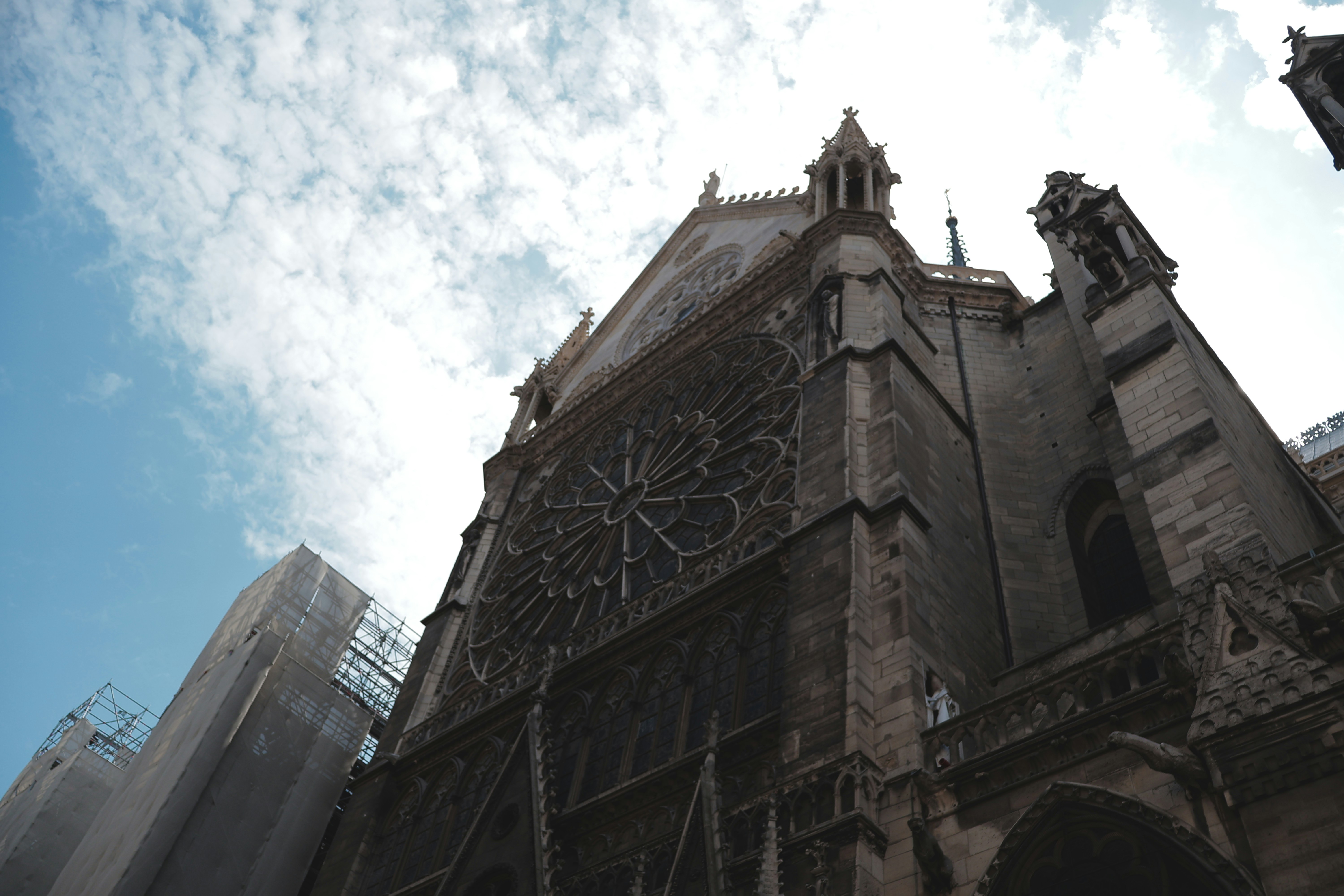 Cathedral facade with intricate rose window and spires against a blue sky; architectural details and contrast between old stone and nearby scaffolding.