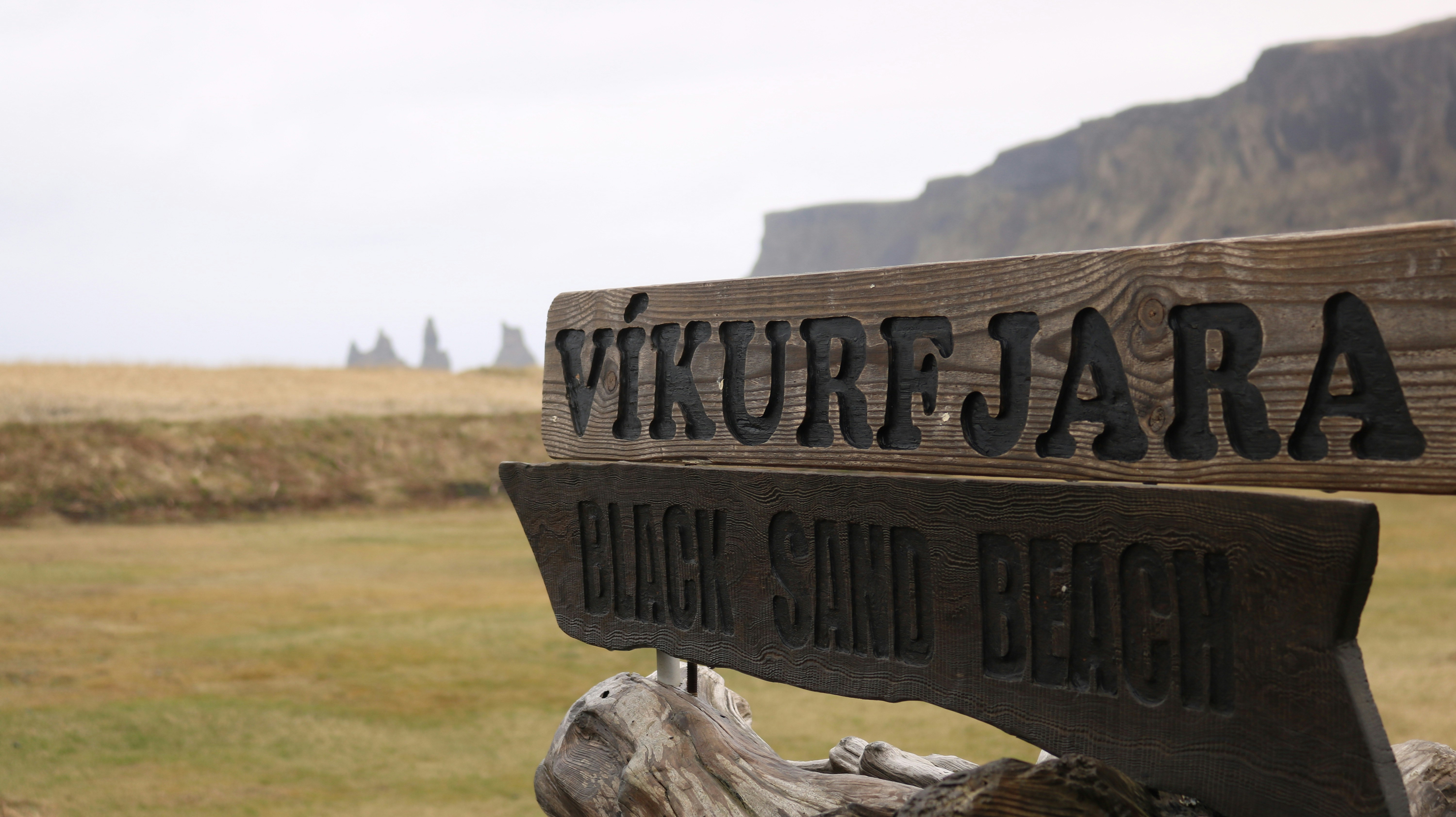 Wooden sign reading 'VIKURFJARA BLACK SAND BEACH' stands in foreground with distant rocky coastline and grassy plain; sign is the focal element of this landscape scene.