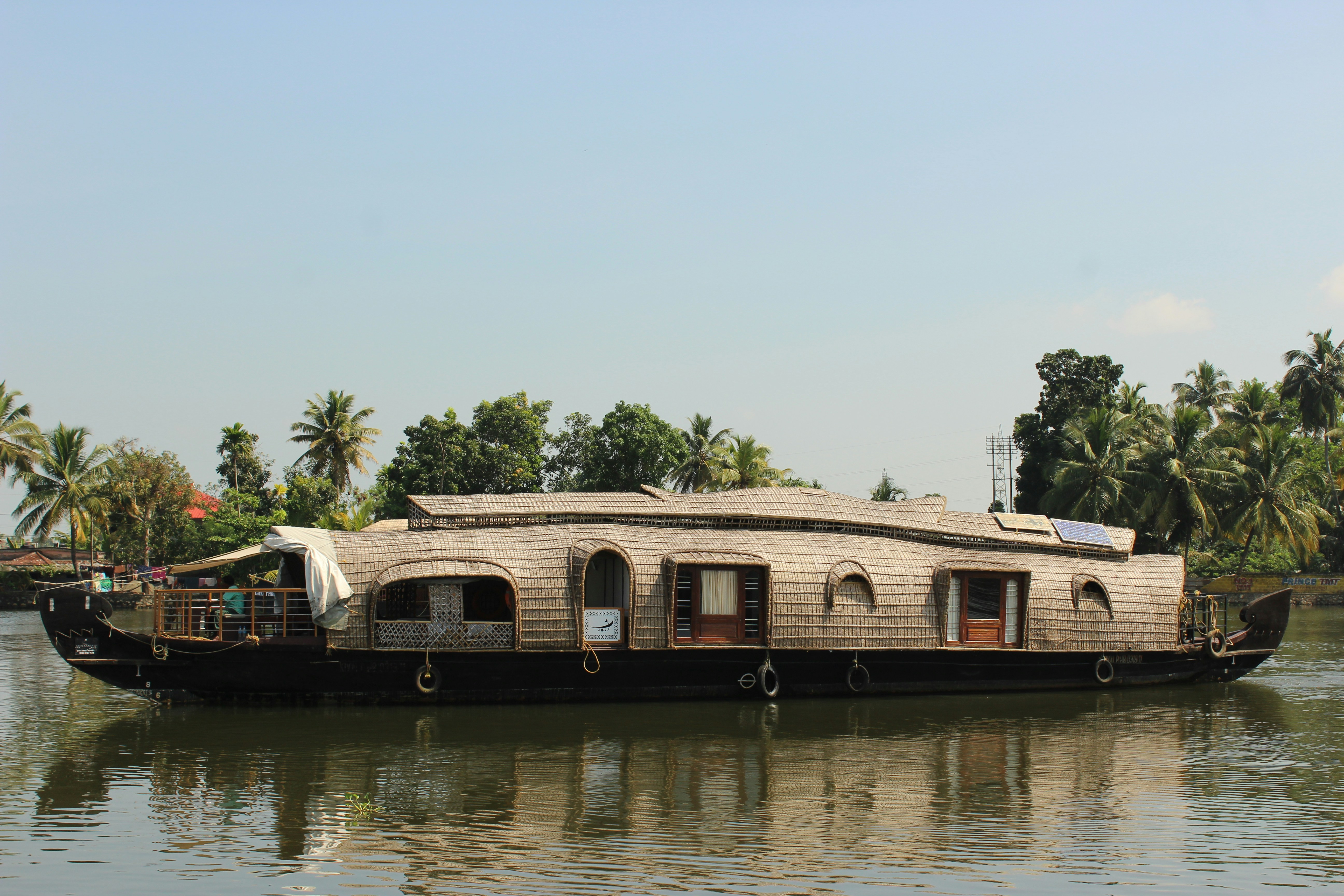 A long houseboat floats on calm river waters with palm trees and a clear sky in the background.