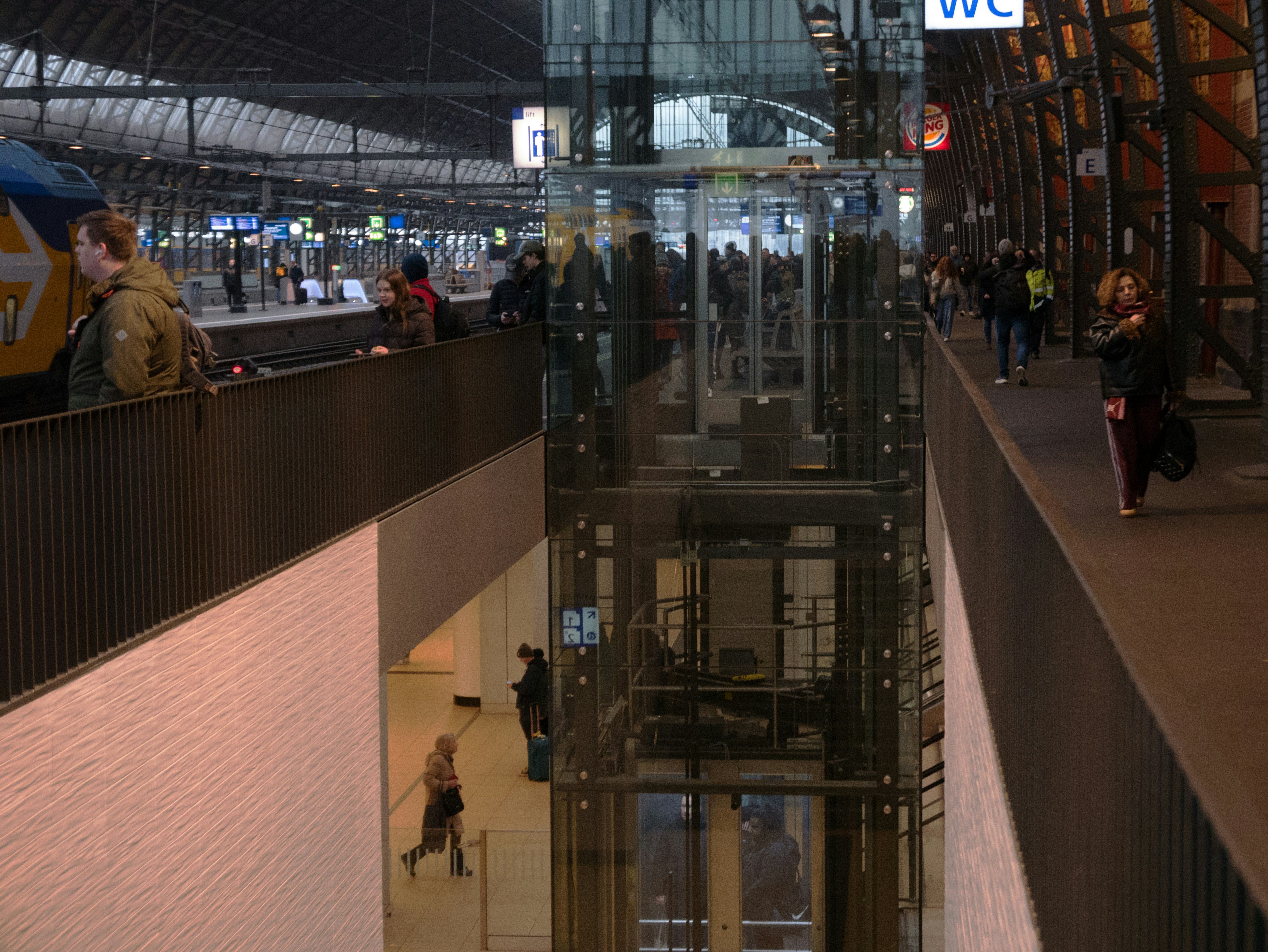 Photo of a glass and transparent elevator shaft at the Central Station Amsterdam between two levens platforms with people, waiting for the train; free image of Dutch street photography in high resolutions, for poster or wallpaper. // Foto van een glazen liftschacht op het Centraal Station Amsterdam, op het perron onder de grote oude stationskap met wachtende mensen; gratis download foto - afbeelding van straatfotografie in Nederland in hoge resolutie, voor poster of wallpaper.
