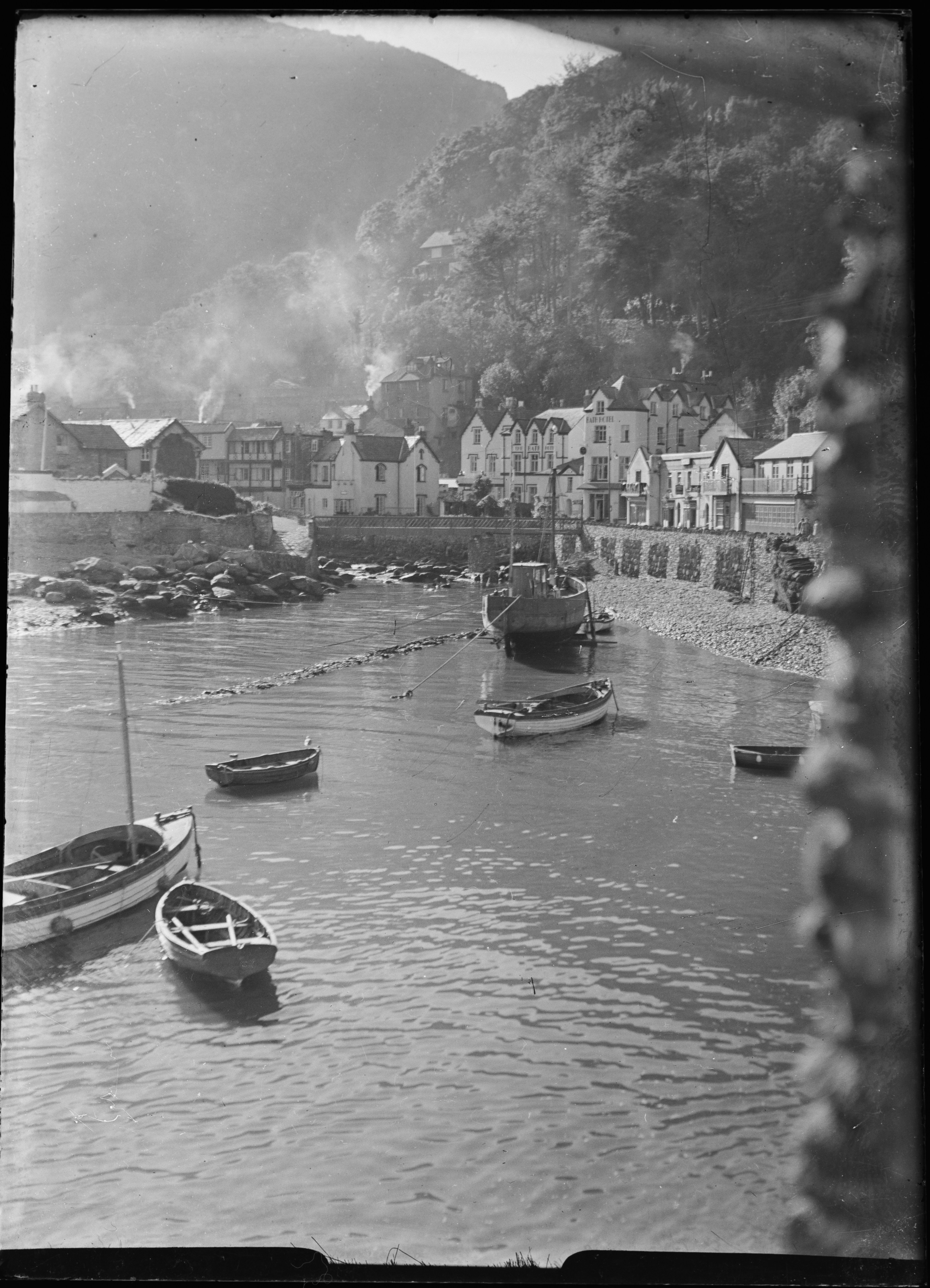 Harbor scene with small boats anchored in a calm inlet, row of houses and hillside in the background; a misty, early-80s feel with muted tones. Photograph captures a coastal village after a calm day; black-and-white medium format texture.