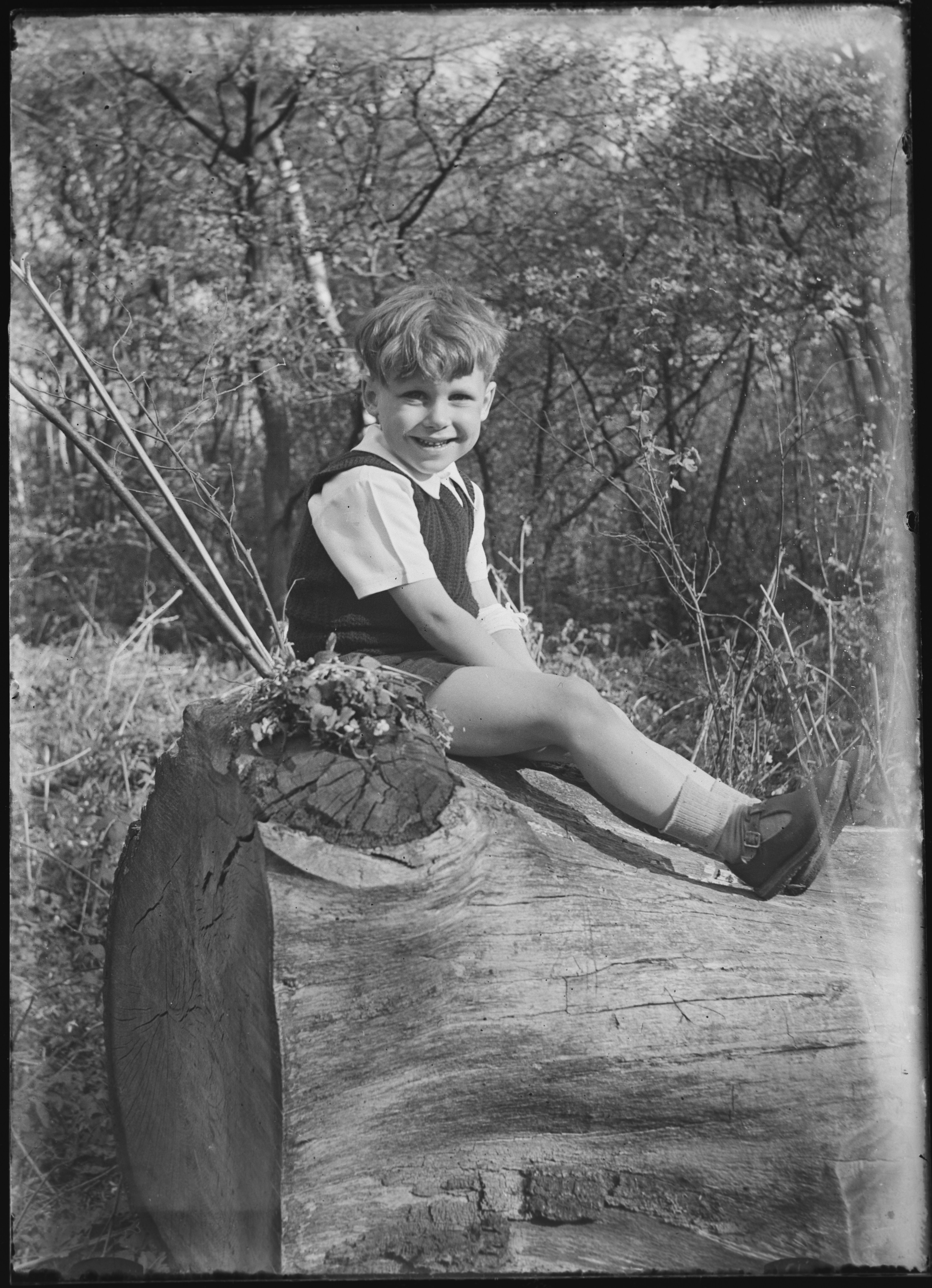 Young boy sits atop a fallen log in a sunlit woodland, smiling toward the camera; vintage grayscale portrait with natural surroundings.