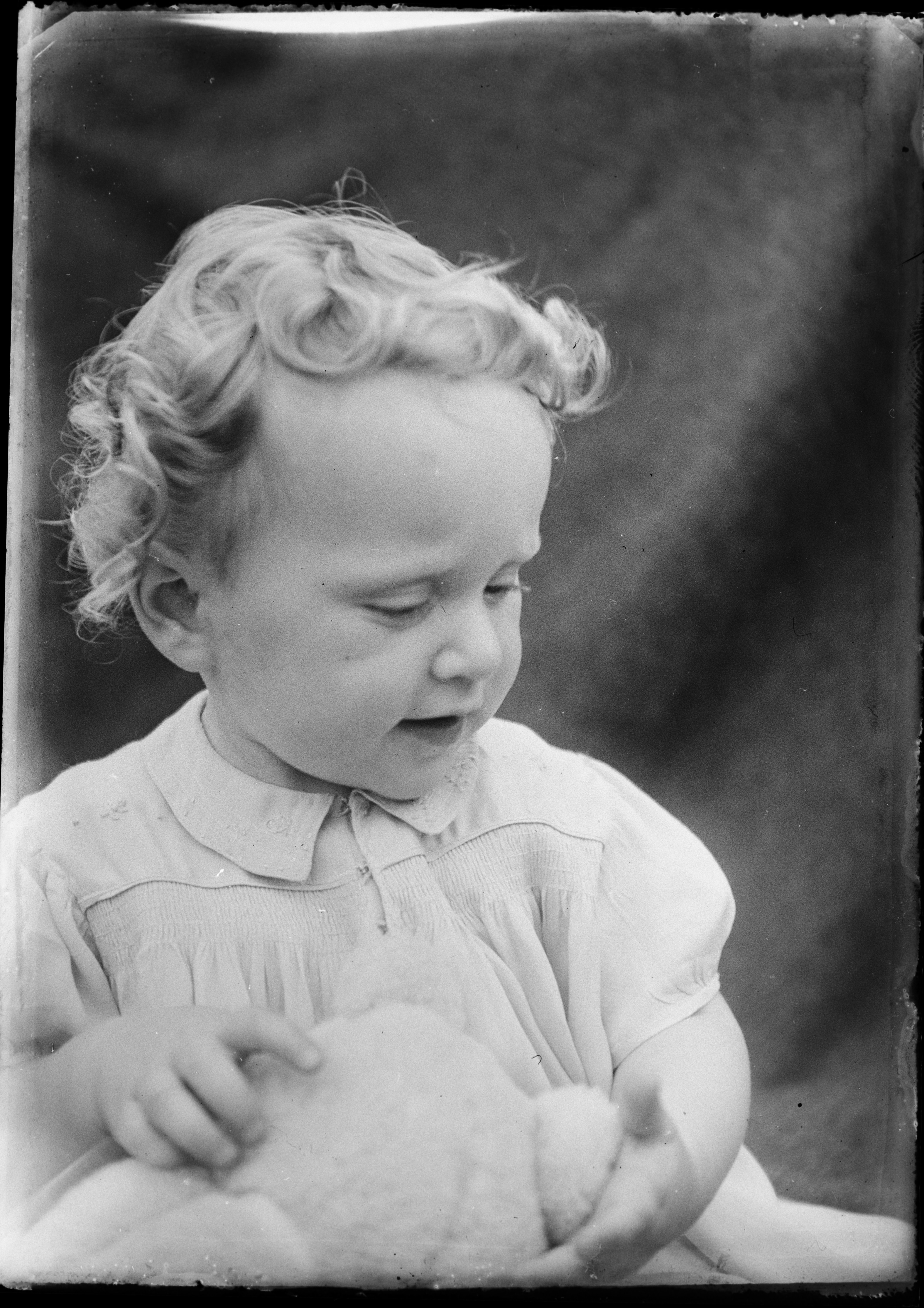 Young child with curly hair gazes downward, holding a soft object in a delicate, vintage-style portrait. A monochrome study focusing on innocence and texture.