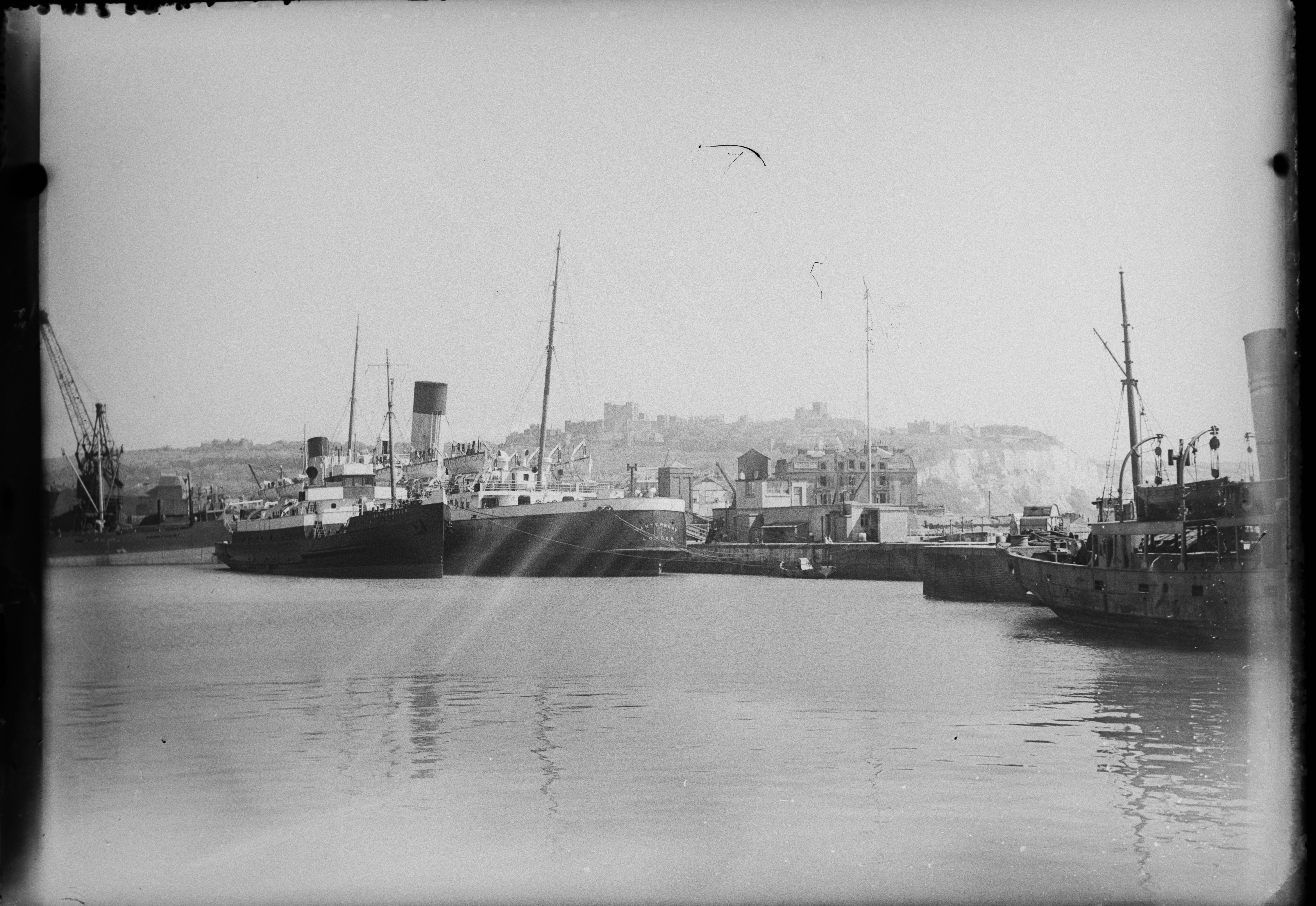 Sunlit harbor with a row of early 20th-century warships resting at the quay; calm water reflects the silhouettes, while distant fortifications rise along the shore.