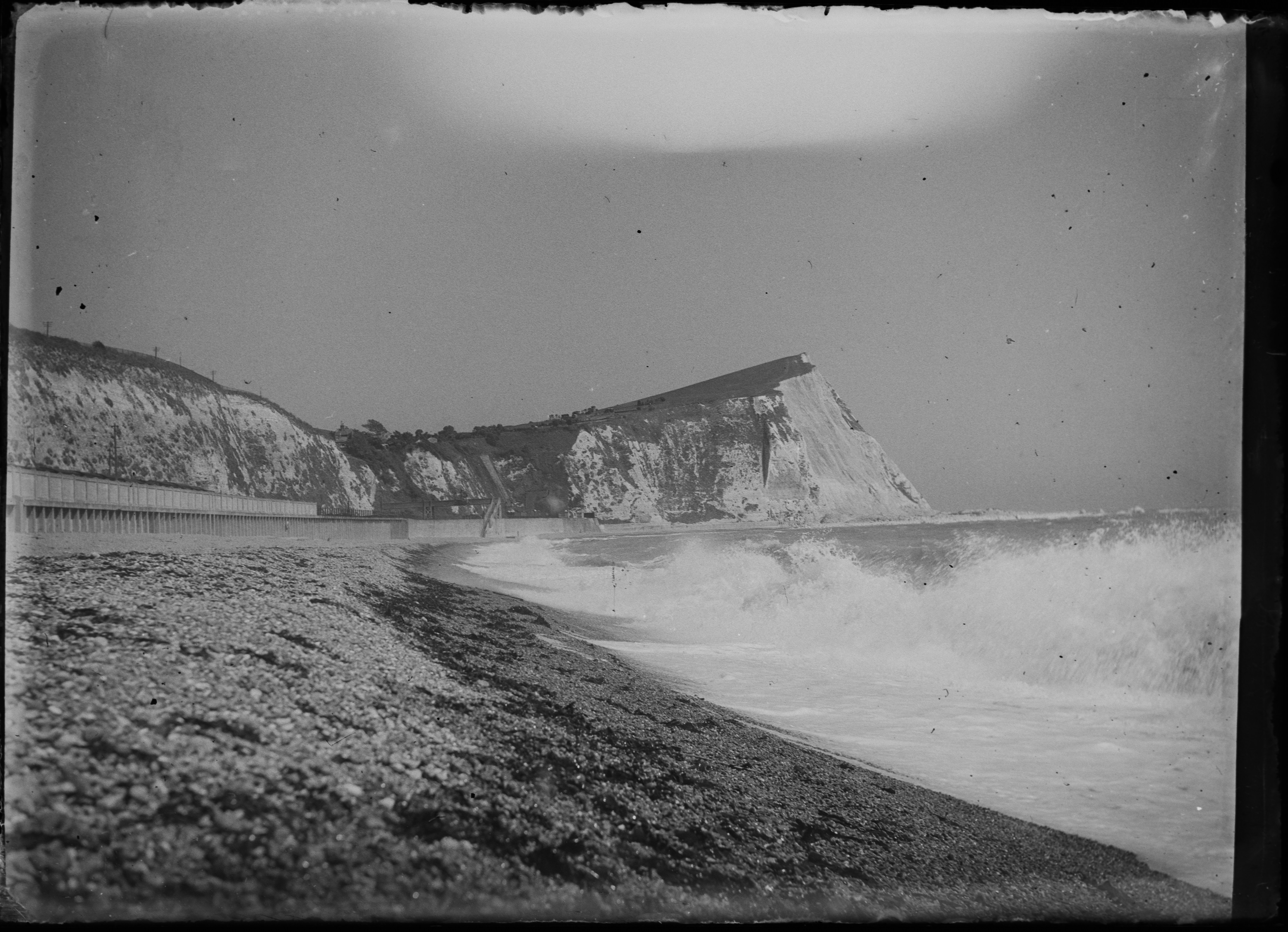 Rocky shoreline with a prominent cliff and surf rolling toward the beach; monochrome capture emphasizing contrast and texture.
