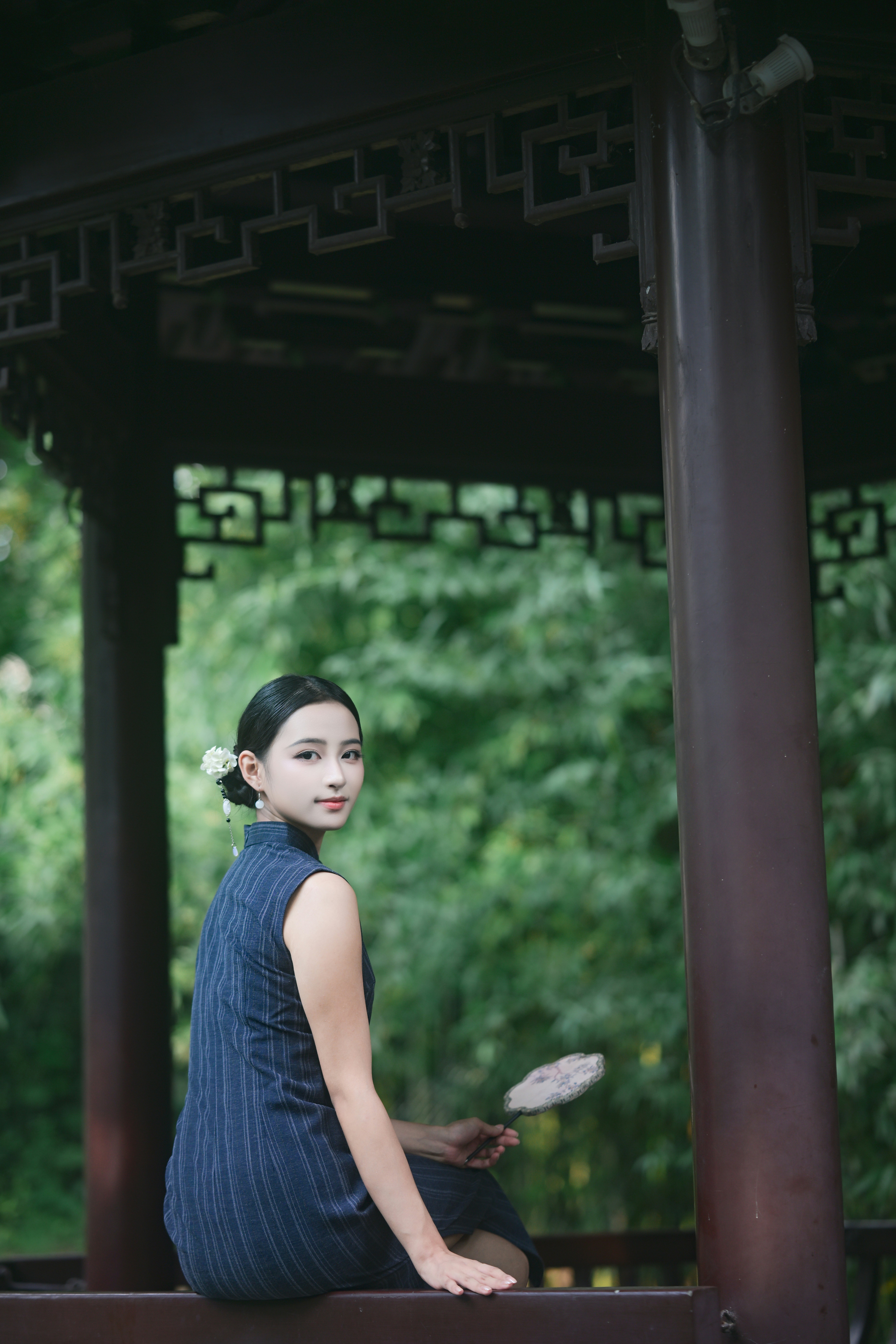 A woman in a blue striped cheongsam holding a fan in a wooden pavilion