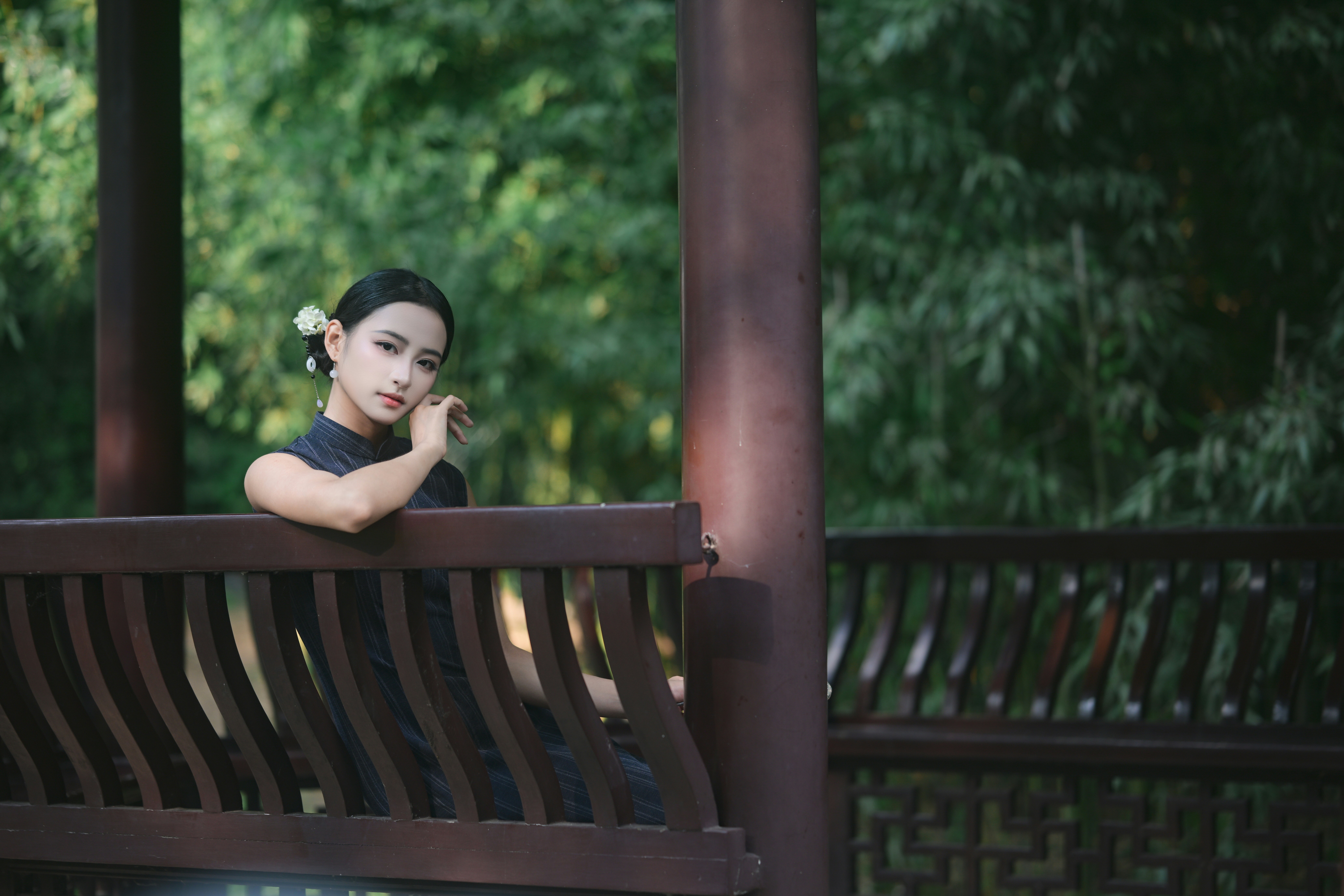 A woman in a dark cheongsam resting in a wooden pavilion with bamboo foliage