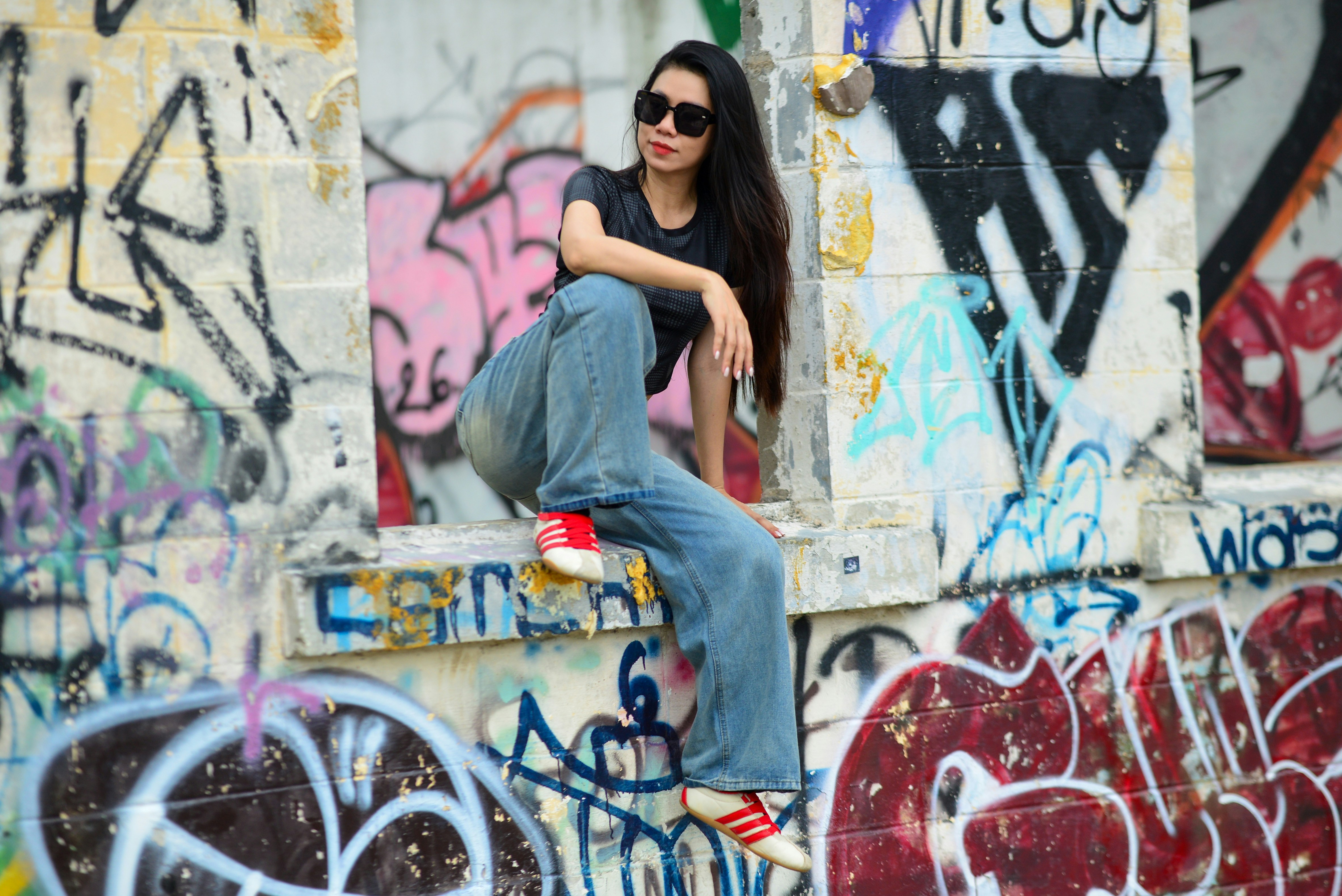 Confident young Asian woman with long dark hair wearing black t-shirt, wide leg denim jeans, red striped sneakers and oversized sunglasses poses seated on a concrete ledge in front of a vibrant graffiti covered wall. Urban street style fashion portrait ideal for youth culture, lifestyle, fashion and city living concepts.